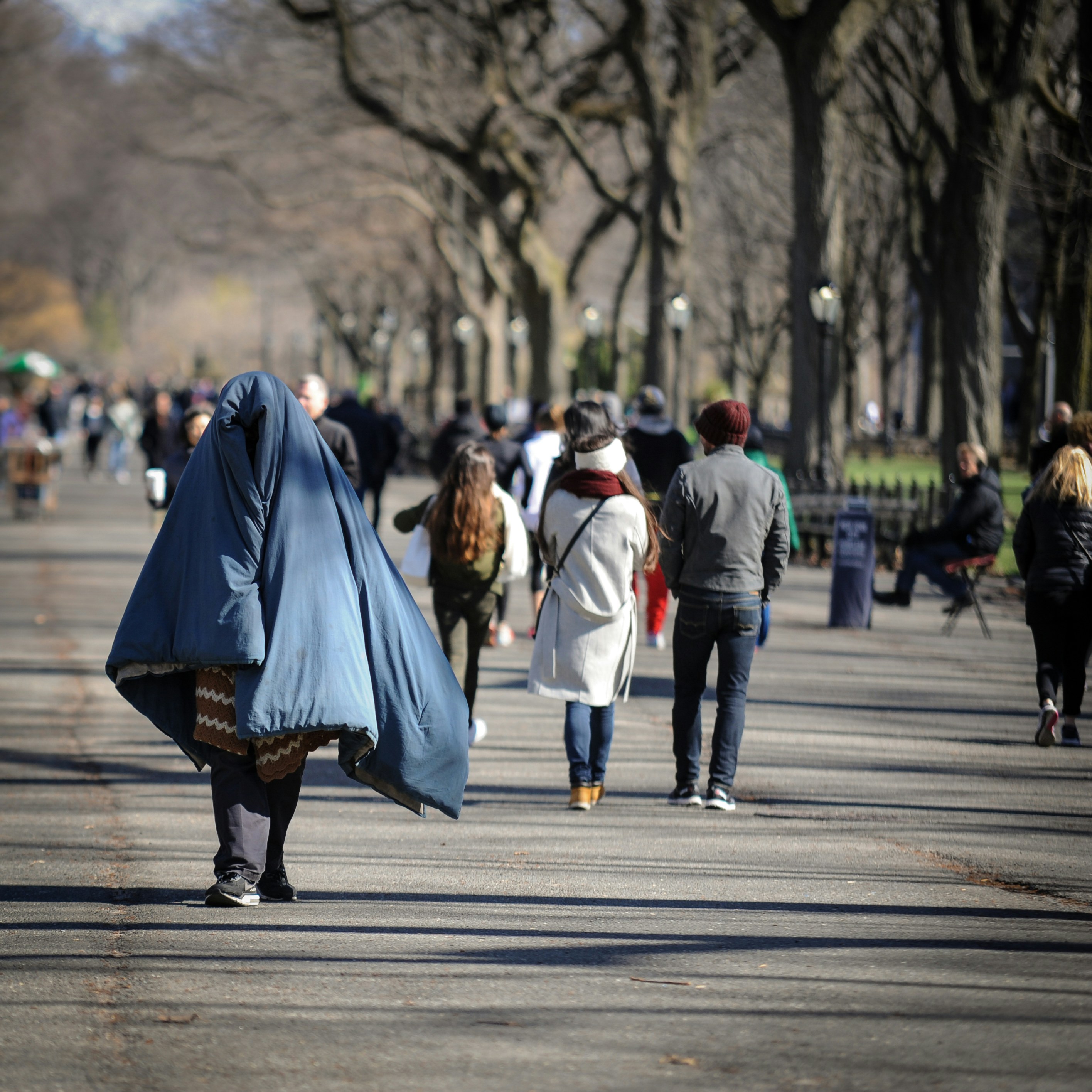A figure draped in a blue blanket walks through a bustling park, contrasting with the lively crowd surrounding them.