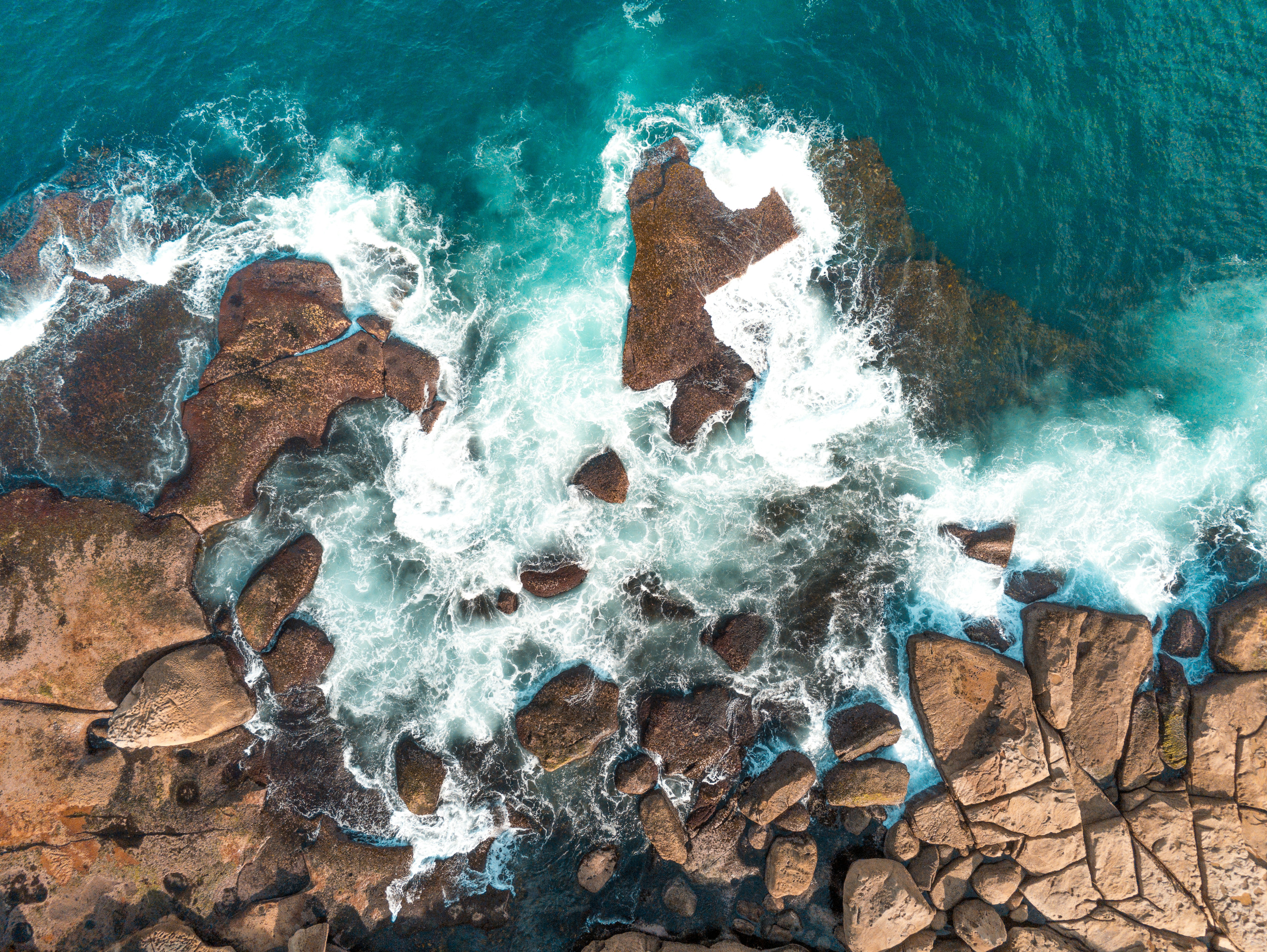 Waves crashing over rugged rocks in turquoise waters from an aerial view.