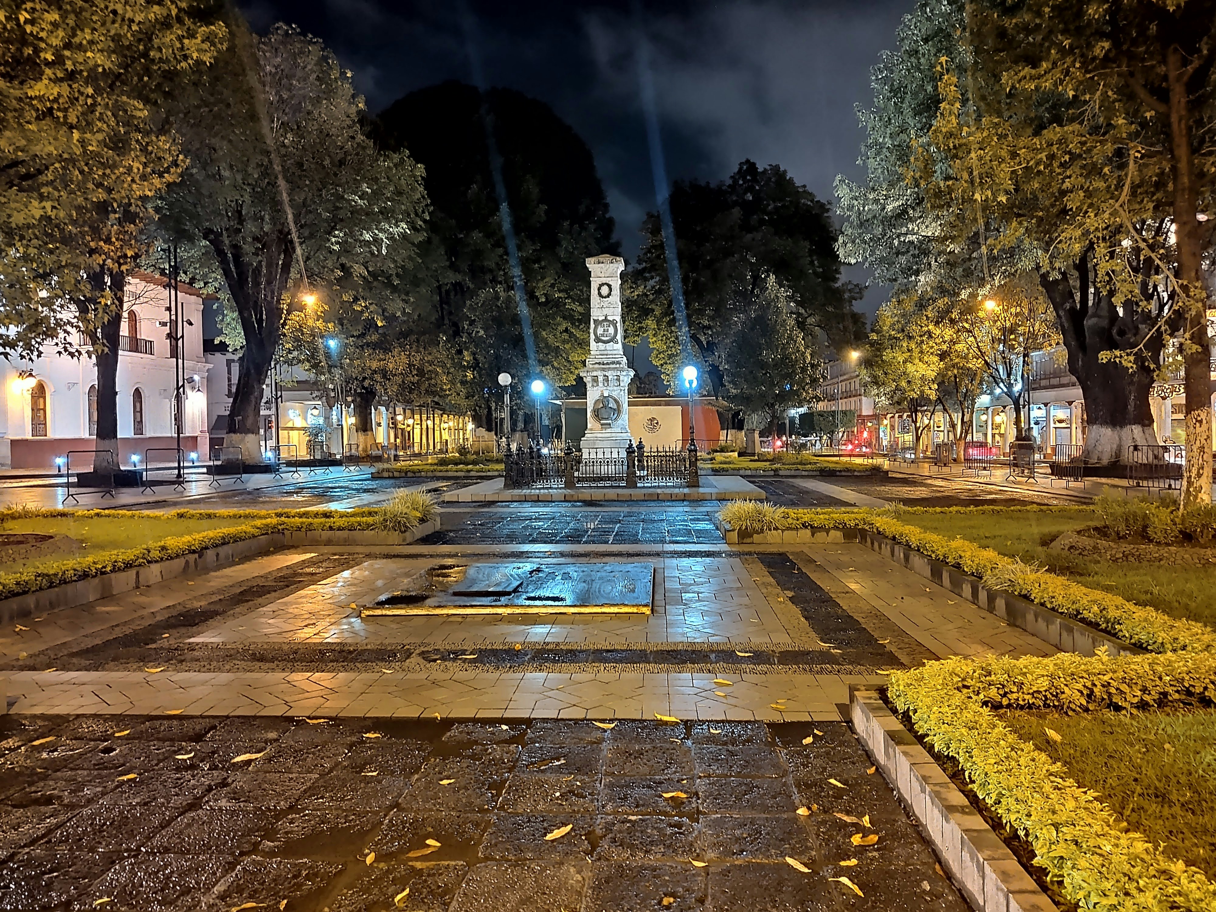Illuminated central plaza in Uruapan with a prominent monument surrounded by trees and trimmed hedges at night.