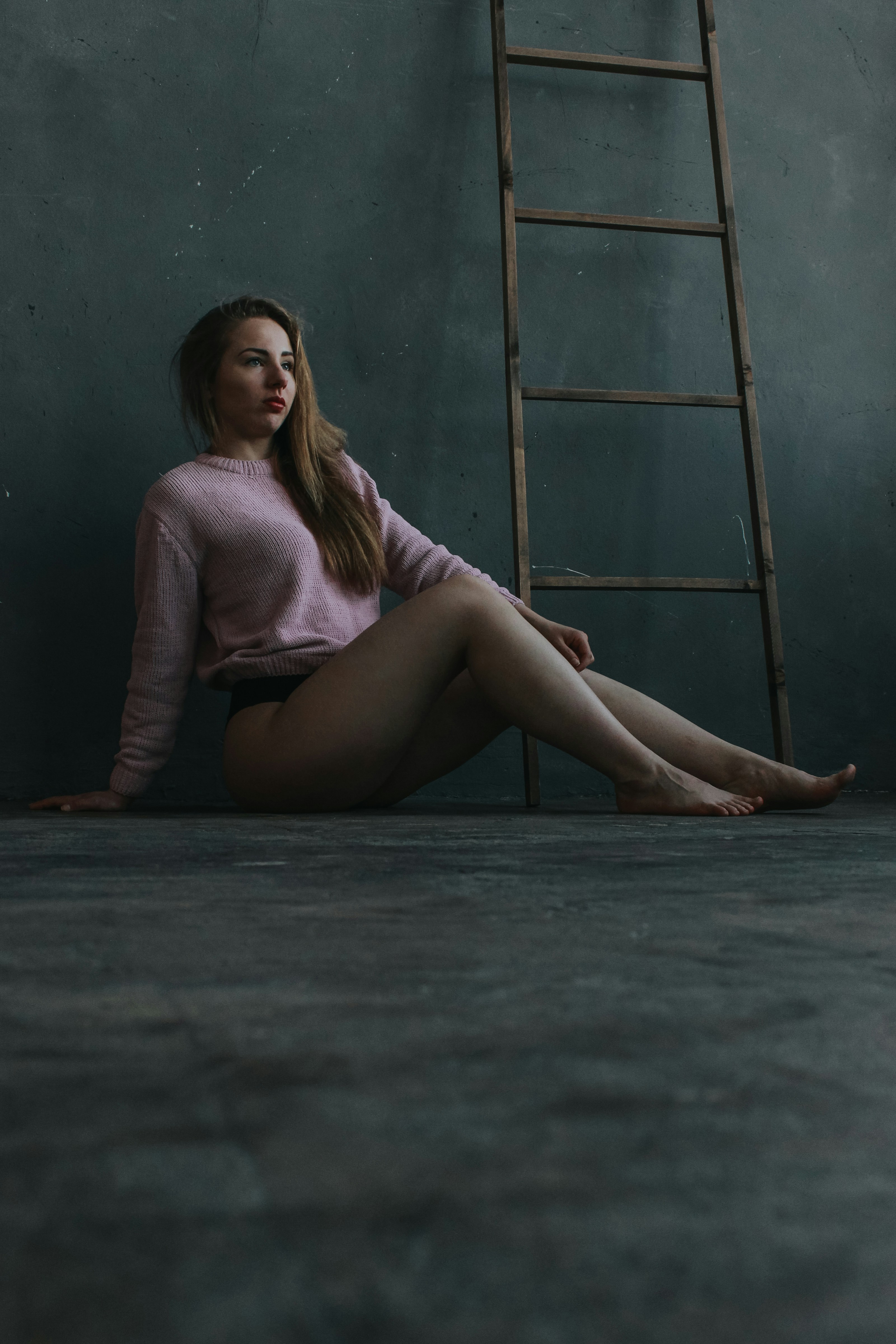 A woman in a soft pink sweater poses gracefully against a textured gray wall, with a rustic wooden ladder nearby.