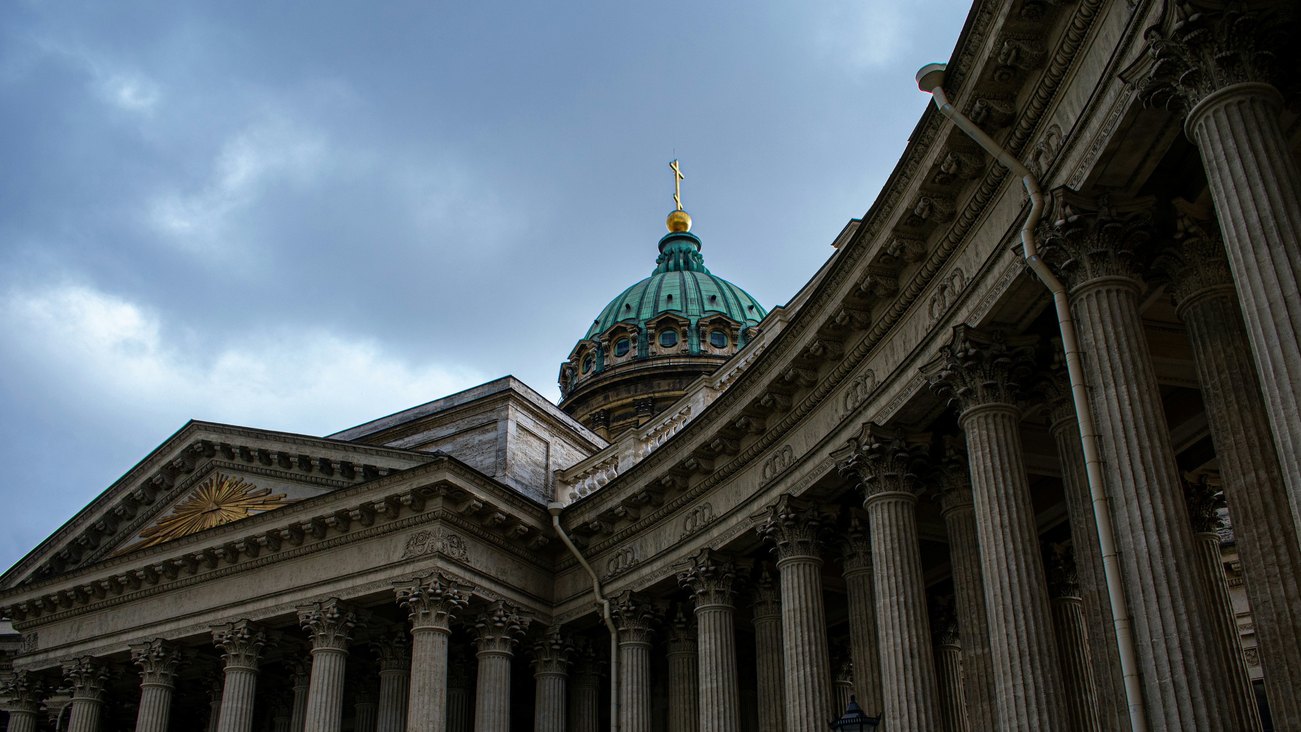 Architectural columns leading to a green-domed structure beneath a cloudy sky.
