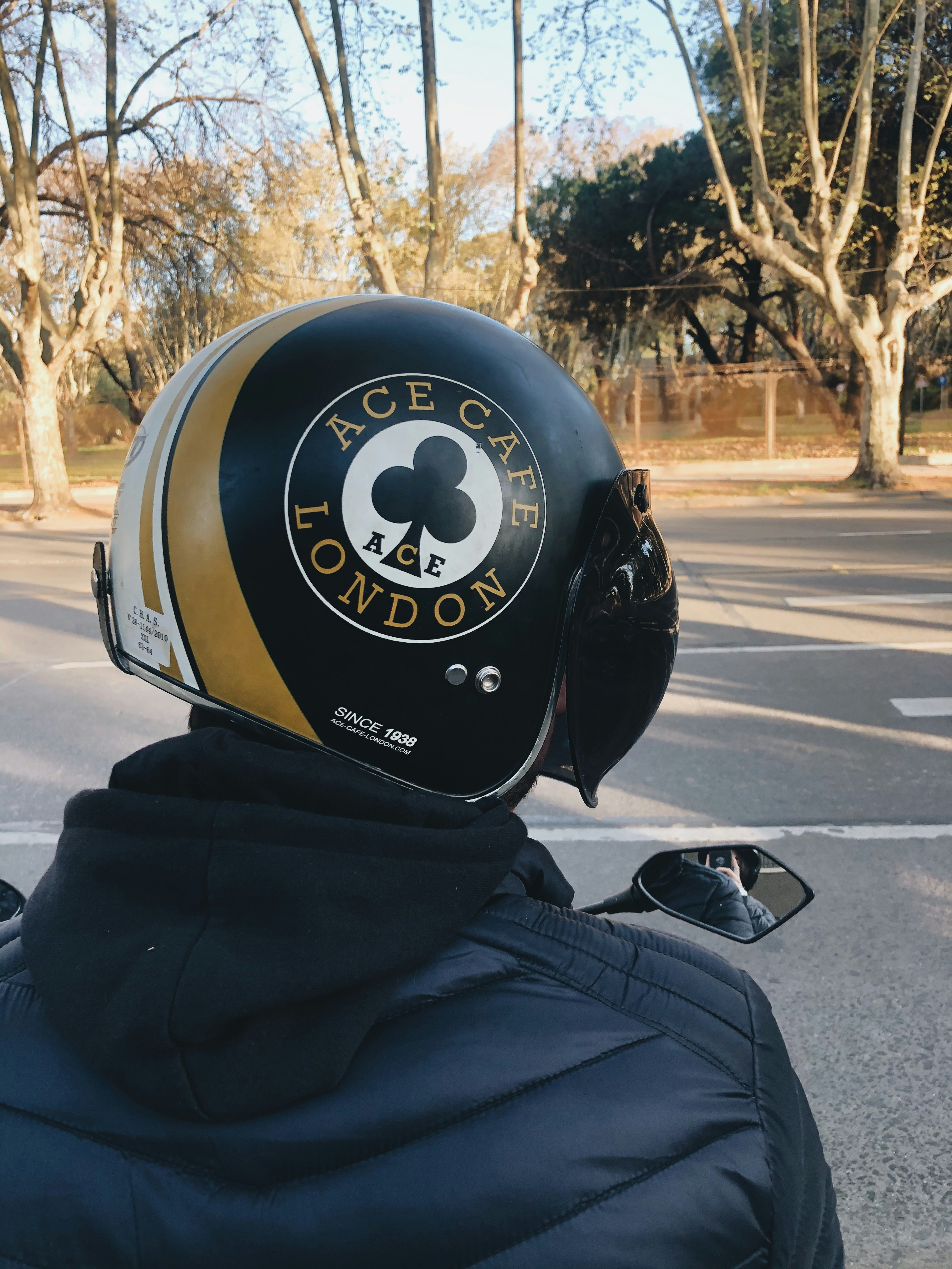 Close-up of a motorcyclist's helmet featuring the Ace Cafe London logo, set against a tree-lined street. The design reflects a classic biker aesthetic.