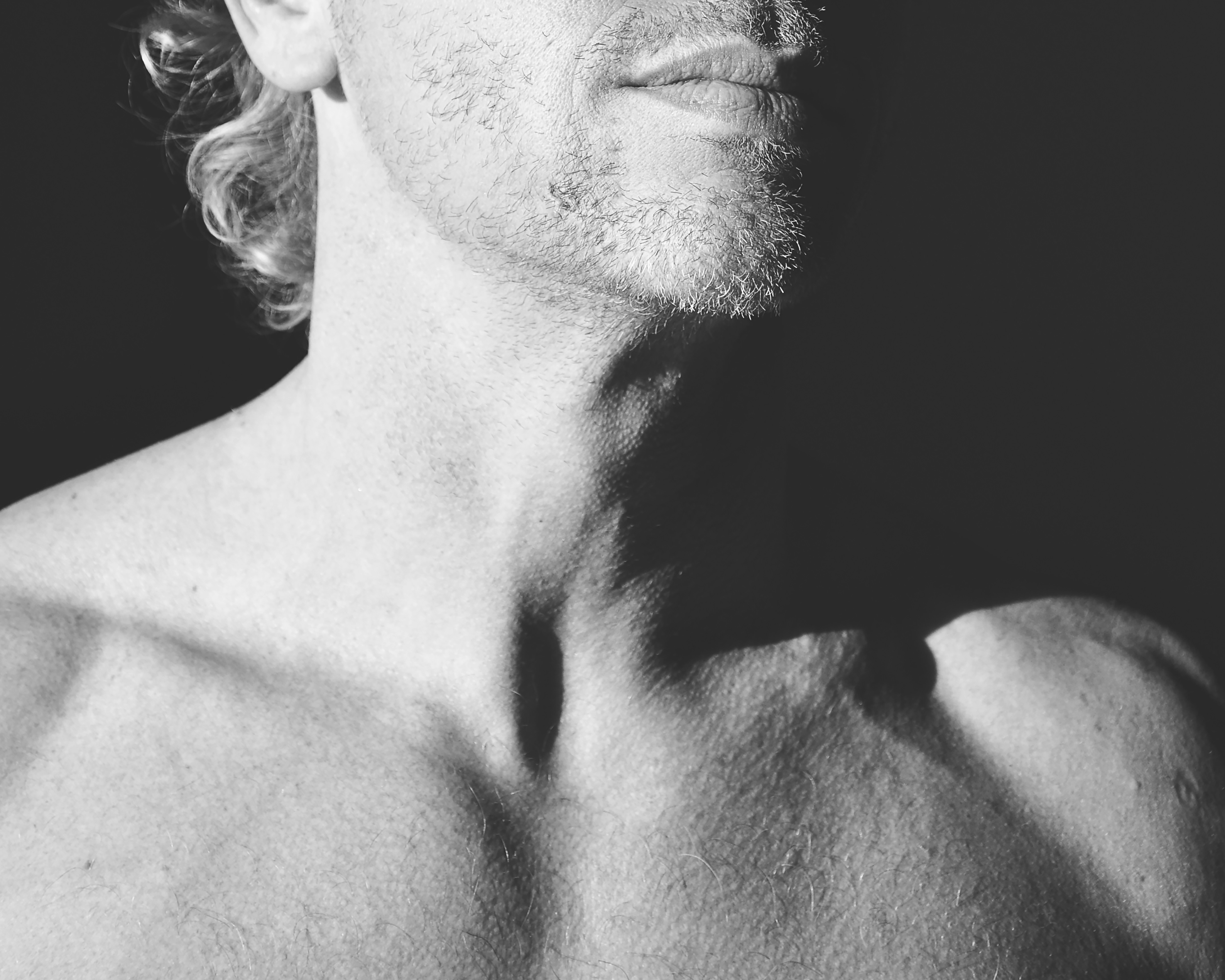 Black and white close-up of a shirtless man's muscular upper body and face partially in shadow.
