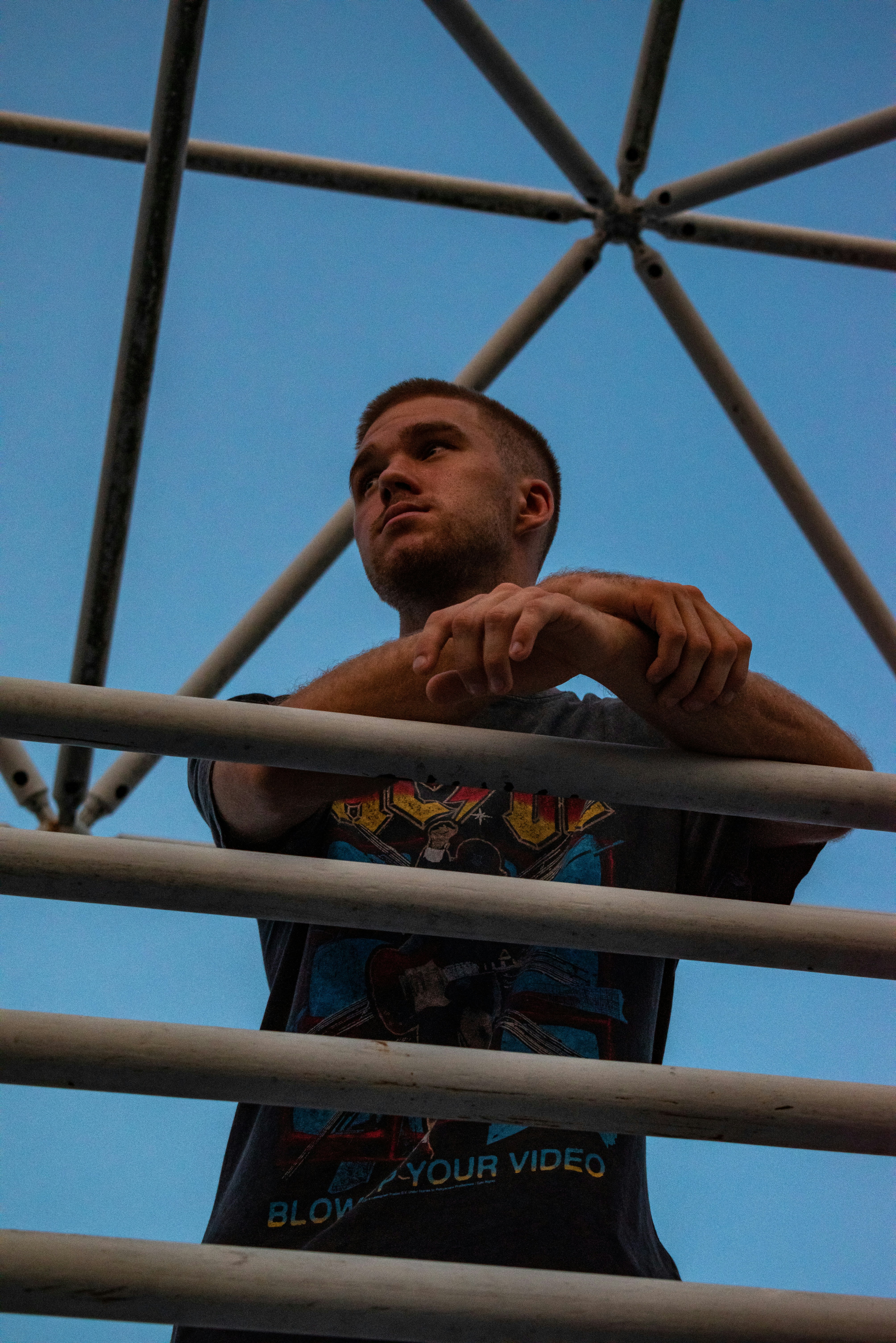Young man leaning on a railing, gazing thoughtfully into the distance, framed by geometric lines against a clear blue sky.