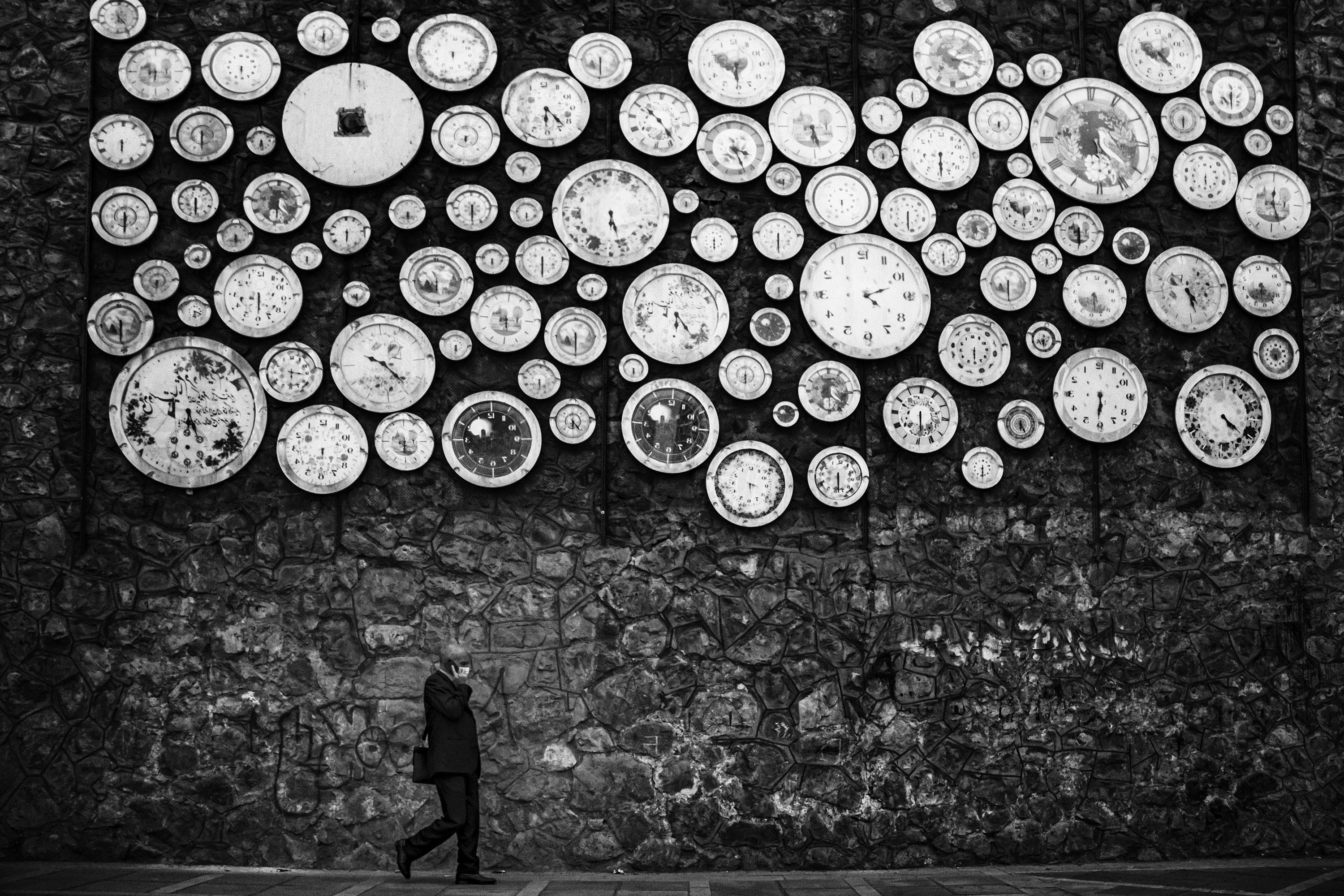 A man walks past an artistic display of clocks mounted on a stone wall, capturing the essence of time in a monochromatic setting.