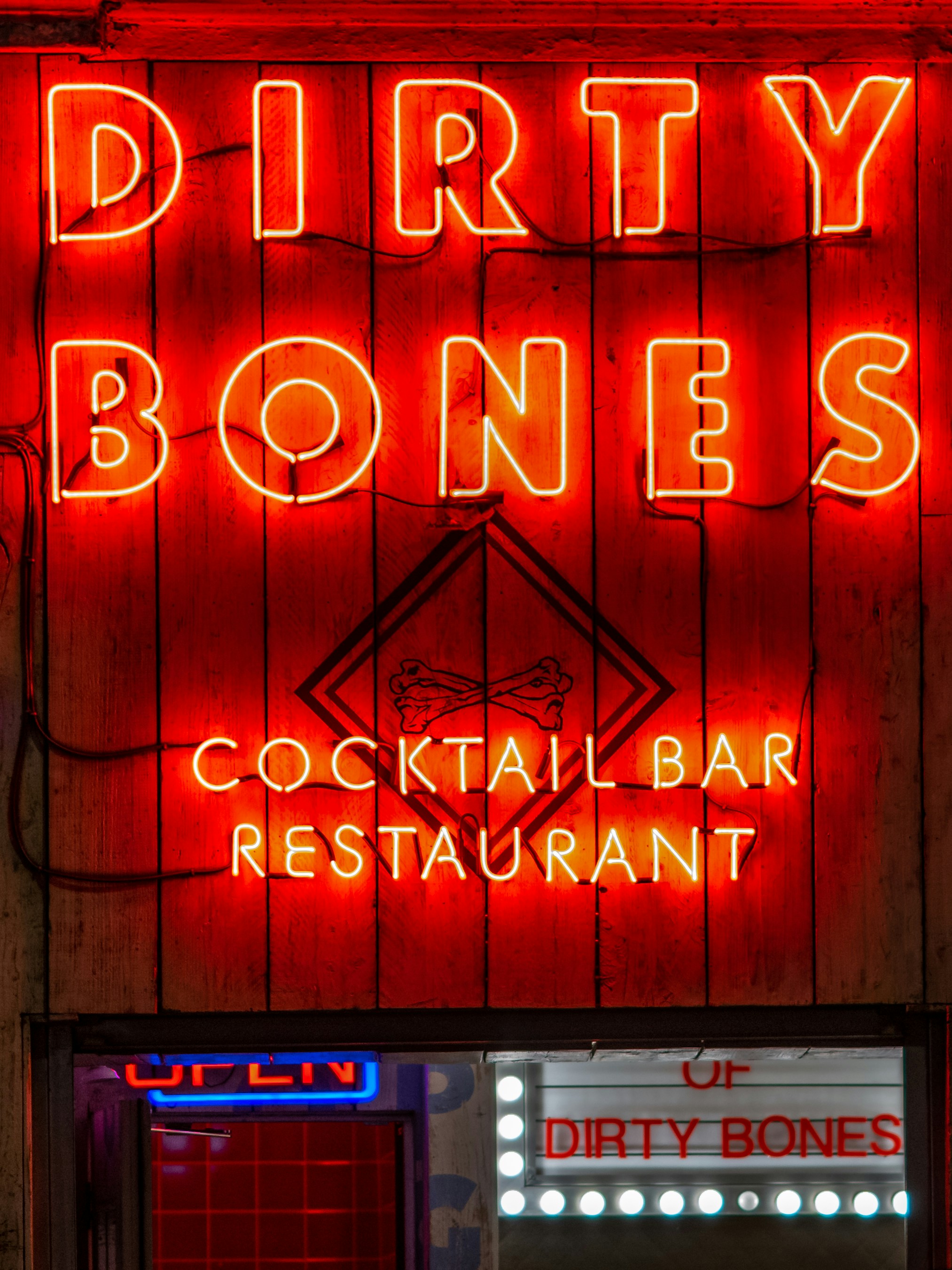 Neon red sign for Dirty Bones Cocktail Bar Restaurant lights up a storefront at night. A diamond-shaped emblem sits beneath the bold lettering.