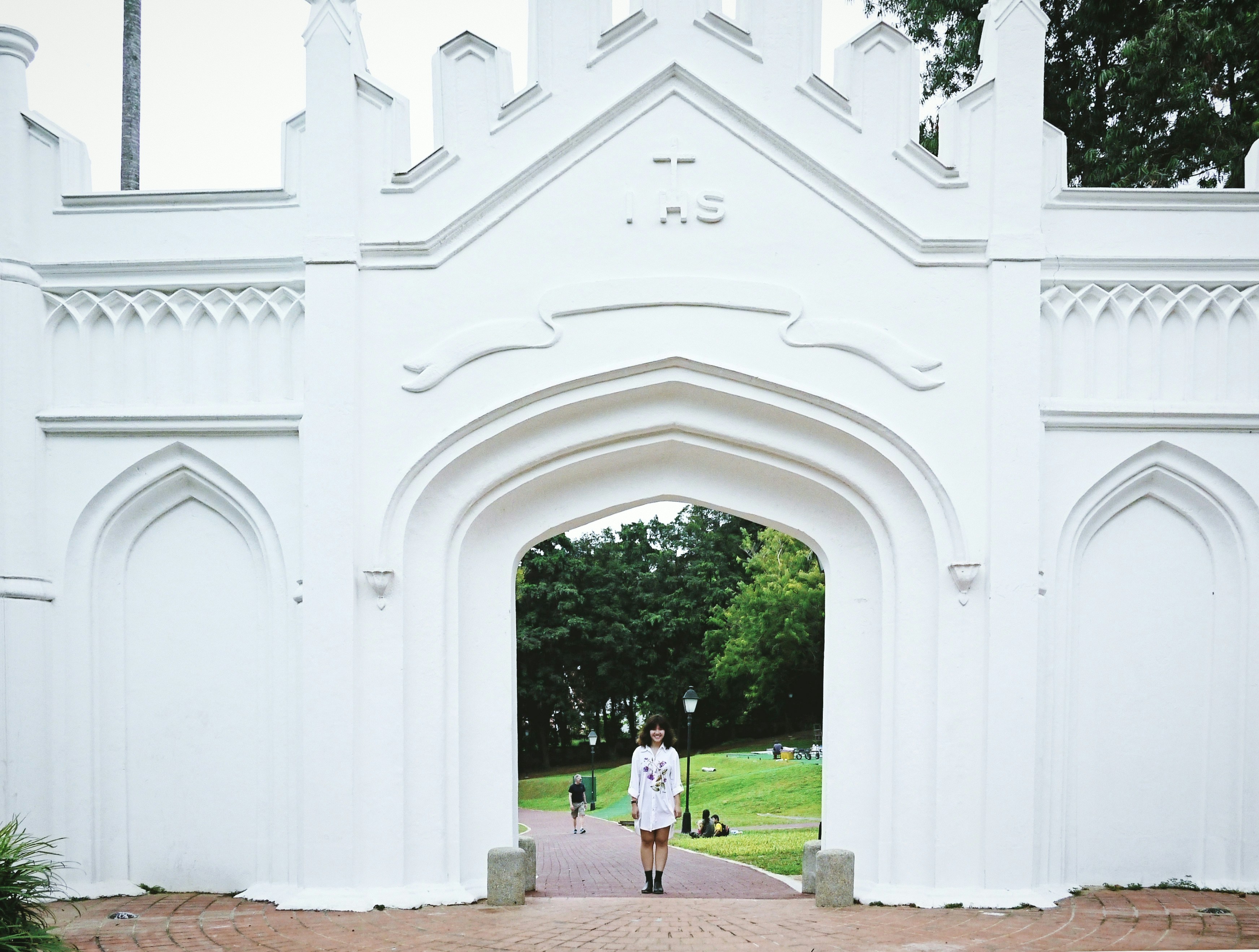 A person stands beneath a grand white archway in a lush park, evoking a sense of tranquility and reflection. The scene captures the harmony between architecture and nature.