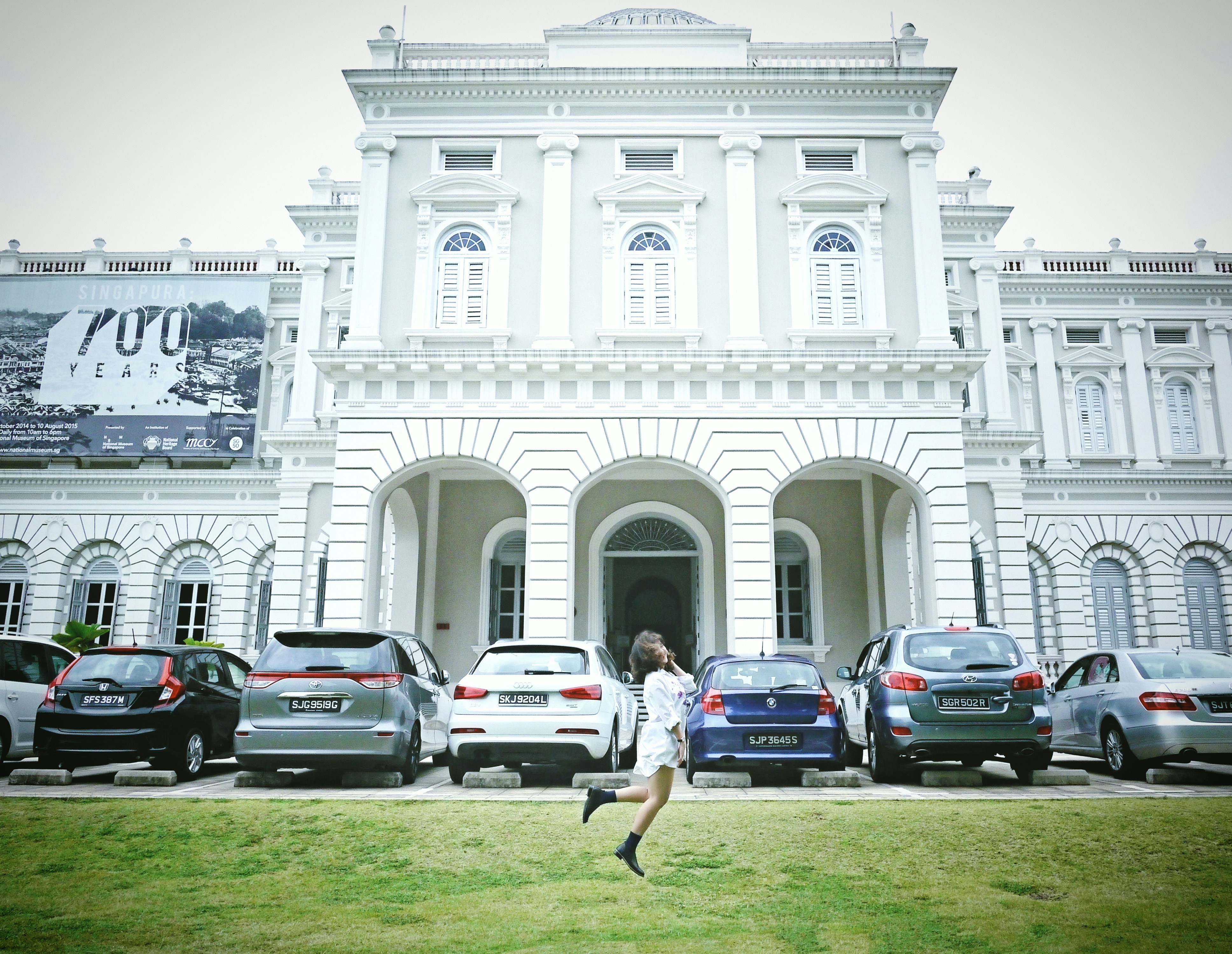 A photograph captures a child sprinting across a manicured lawn in front of a grand Beaux-Arts building, with cars parked along the entrance. The motion contrasts with the formal architectural backdrop.