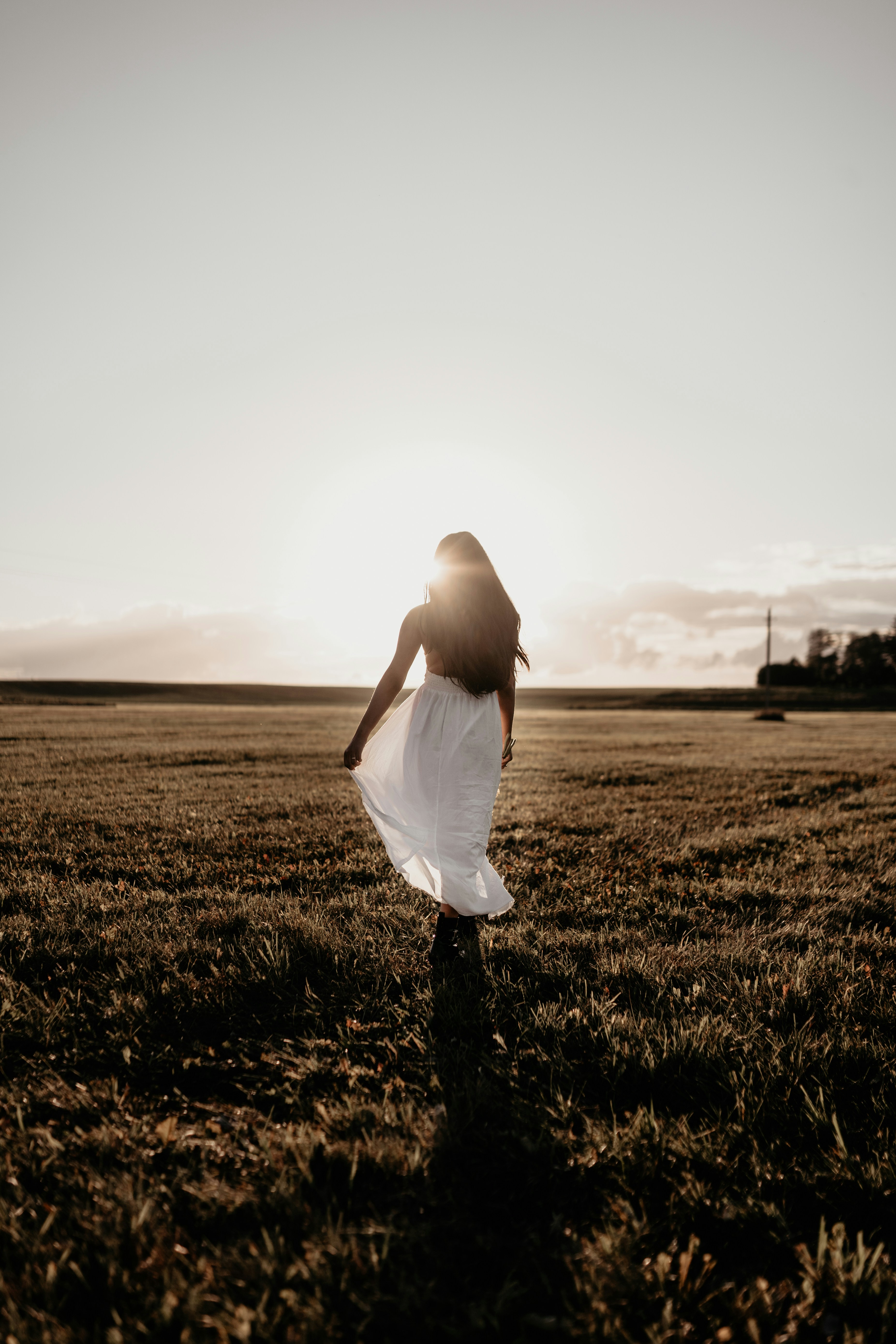 Woman in a flowing white dress walks through a sunlit field, silhouetted against a vibrant sunset. The scene captures a moment of serene solitude.