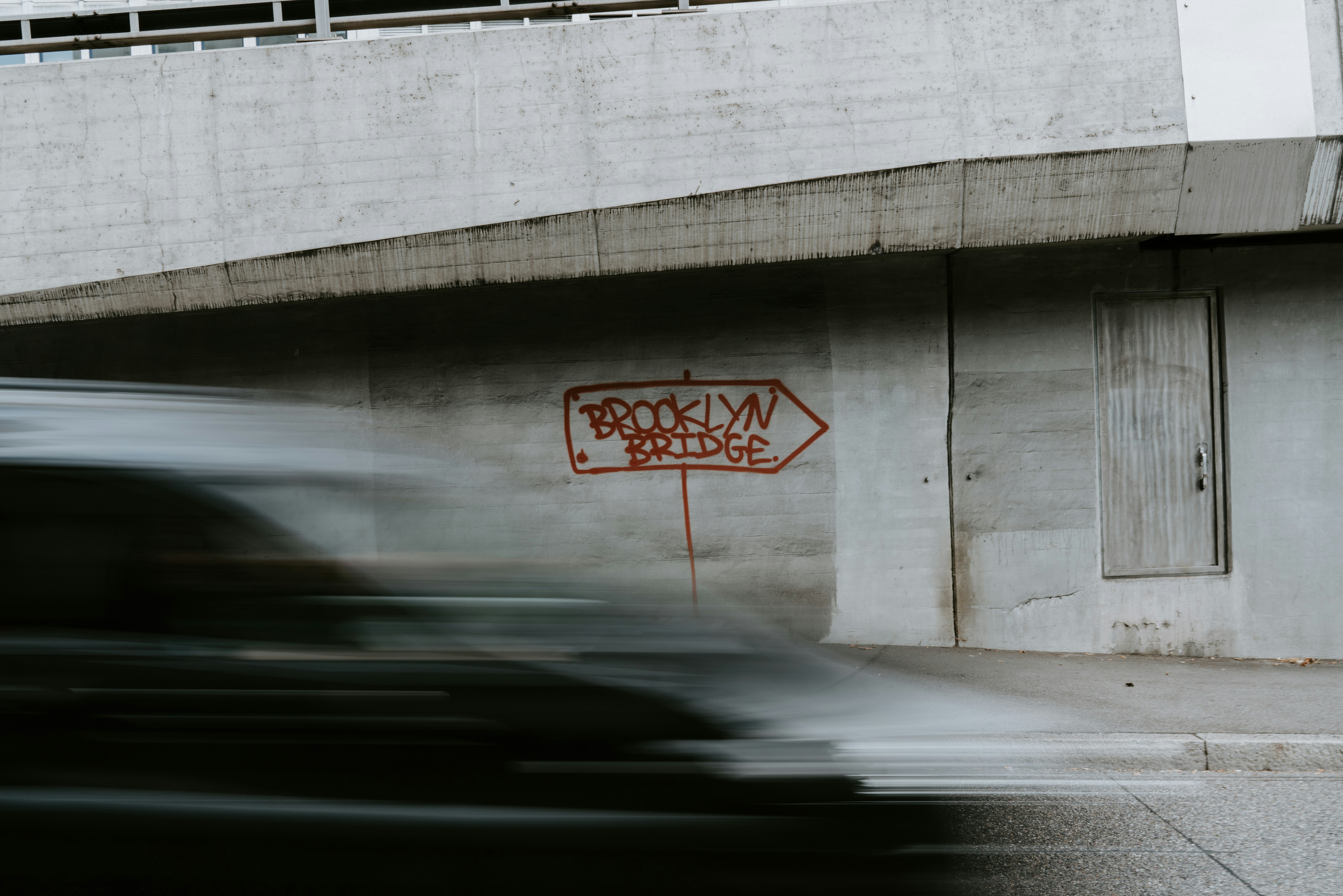 Graffiti indicating 'Brooklyn Bridge' on a concrete wall, with a blurred car passing by, capturing the essence of city life.