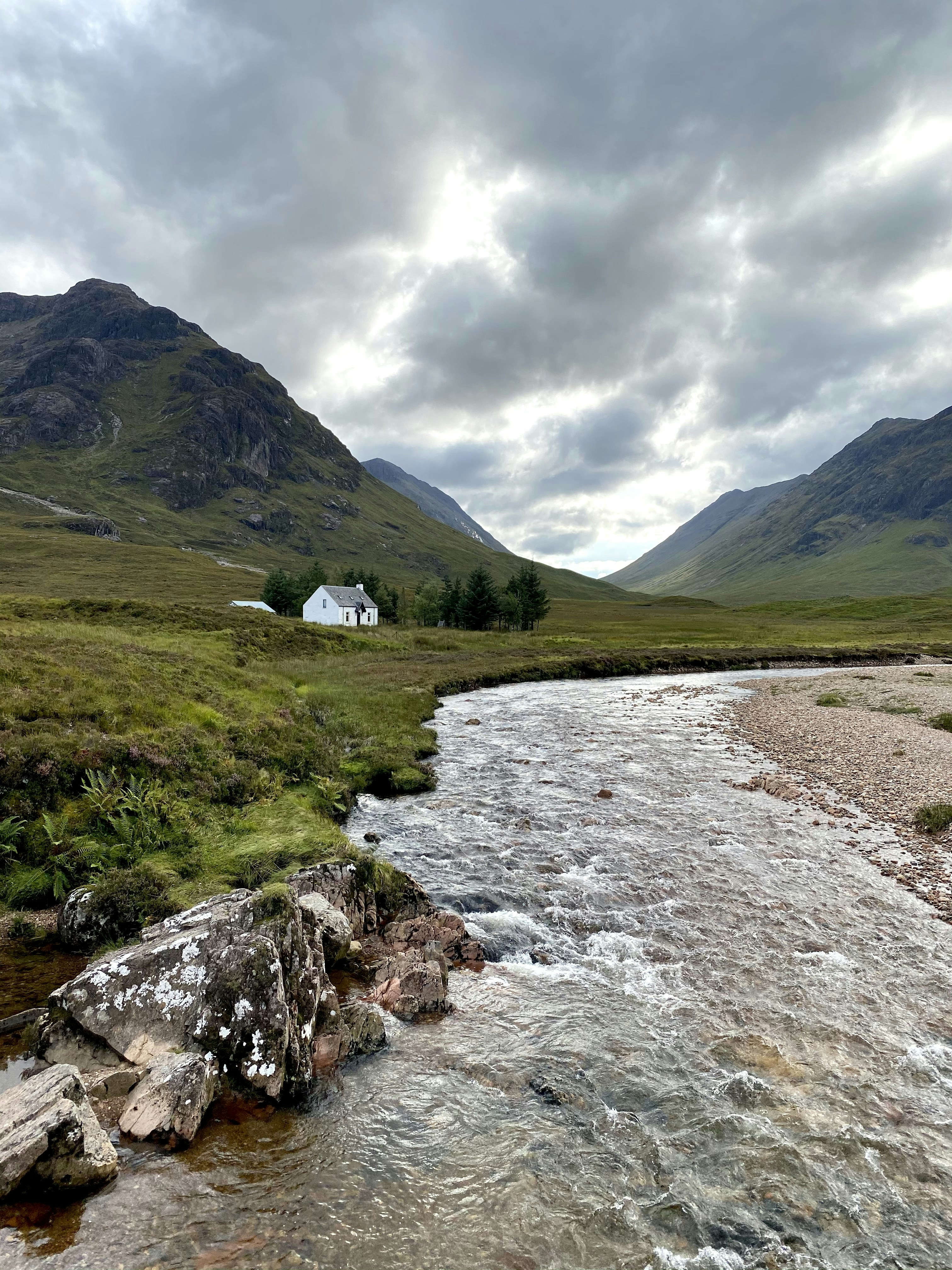 A serene stream meanders through the lush Scottish Highlands, with a quaint white cottage nestled among the trees in the distance. Dramatic mountains rise under a moody sky.