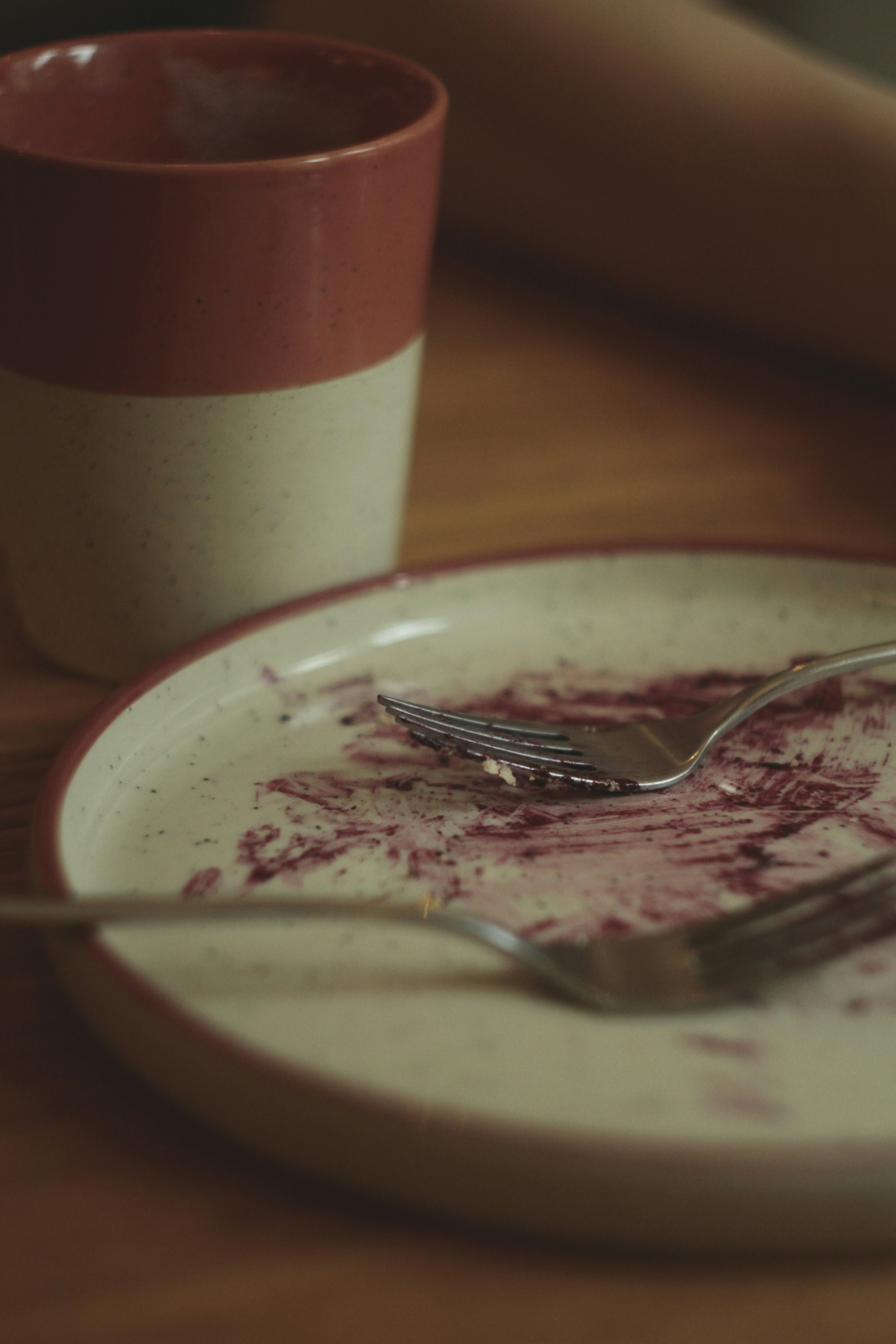 A used plate with traces of food and two forks resting on it, accompanied by a rustic cup, suggesting a recent dining experience.