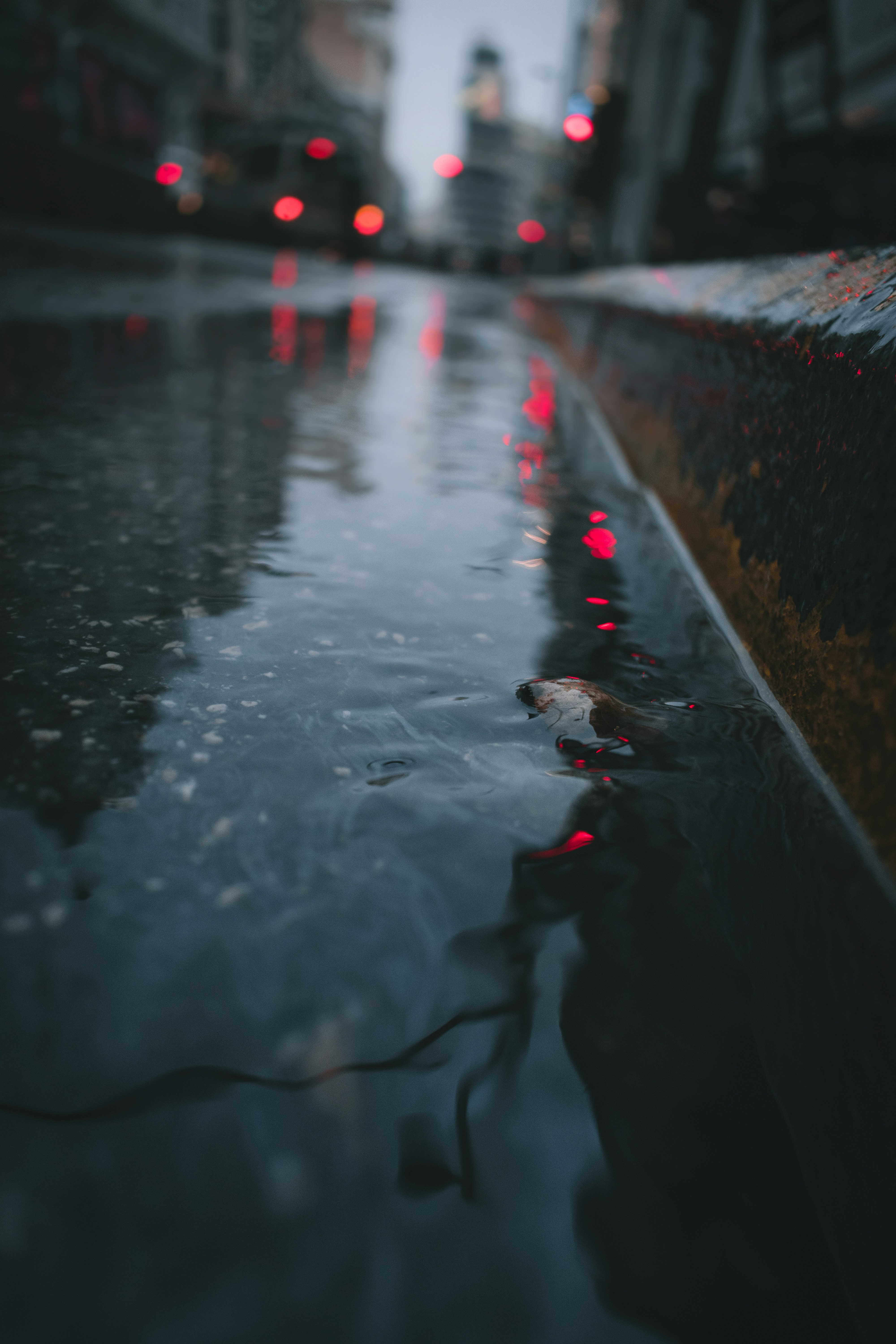 Water pooling in a city street reflects red traffic lights and blurred buildings, creating a moody urban scene.