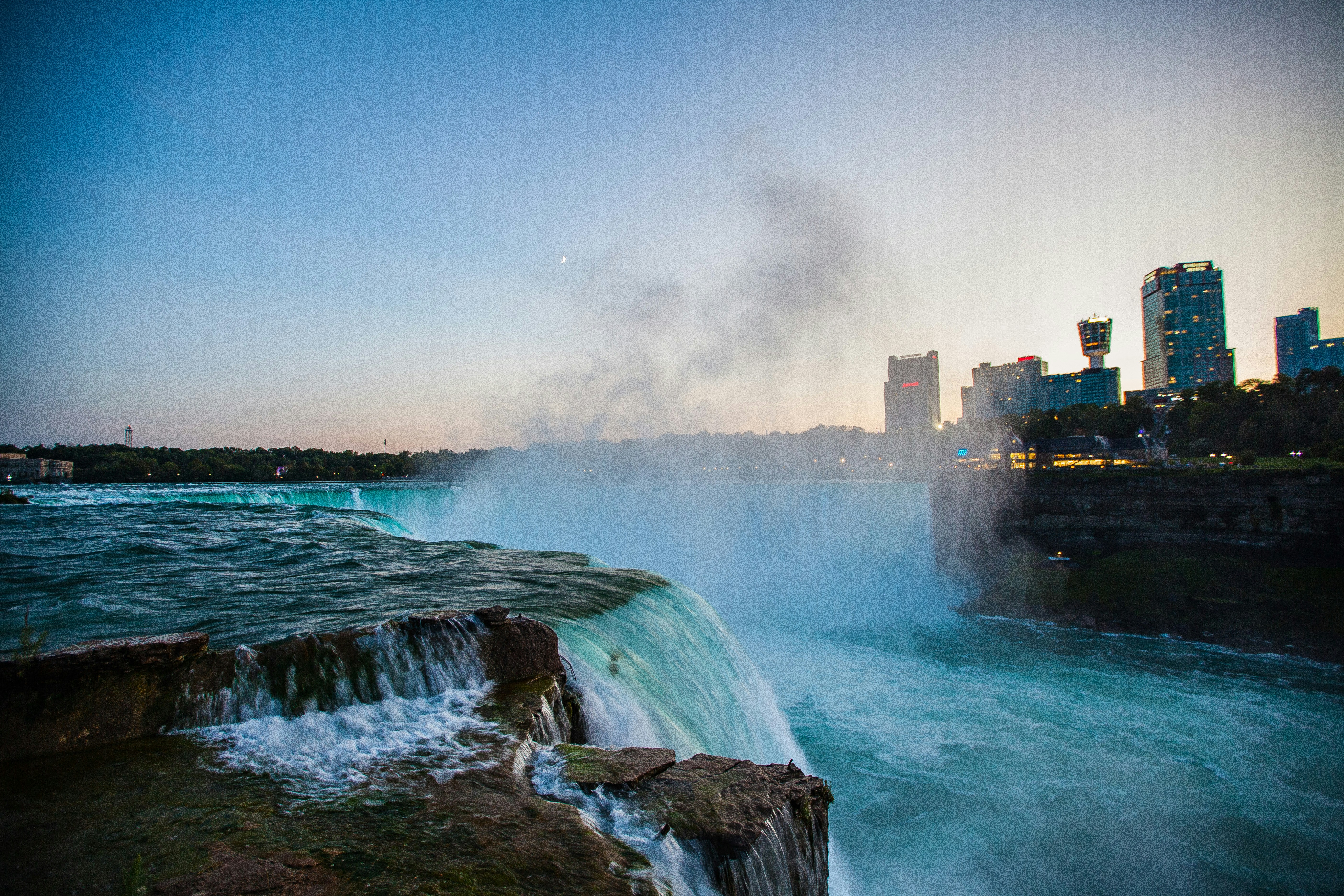Majestic waterfall flowing over rocky ledge with city skyline in the background during twilight.