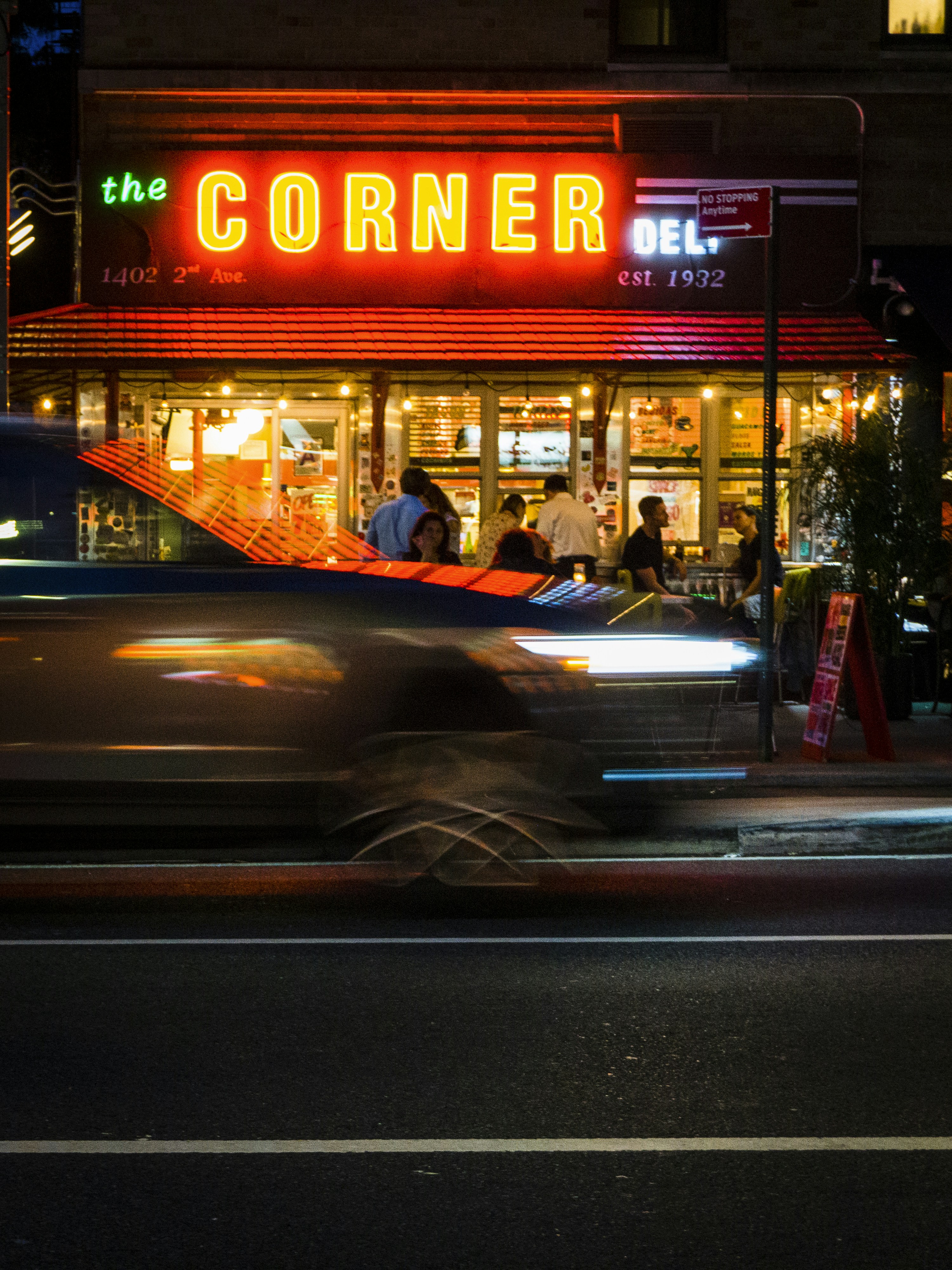 Car blurs past a brightly lit neon restaurant sign in a bustling city street at night.