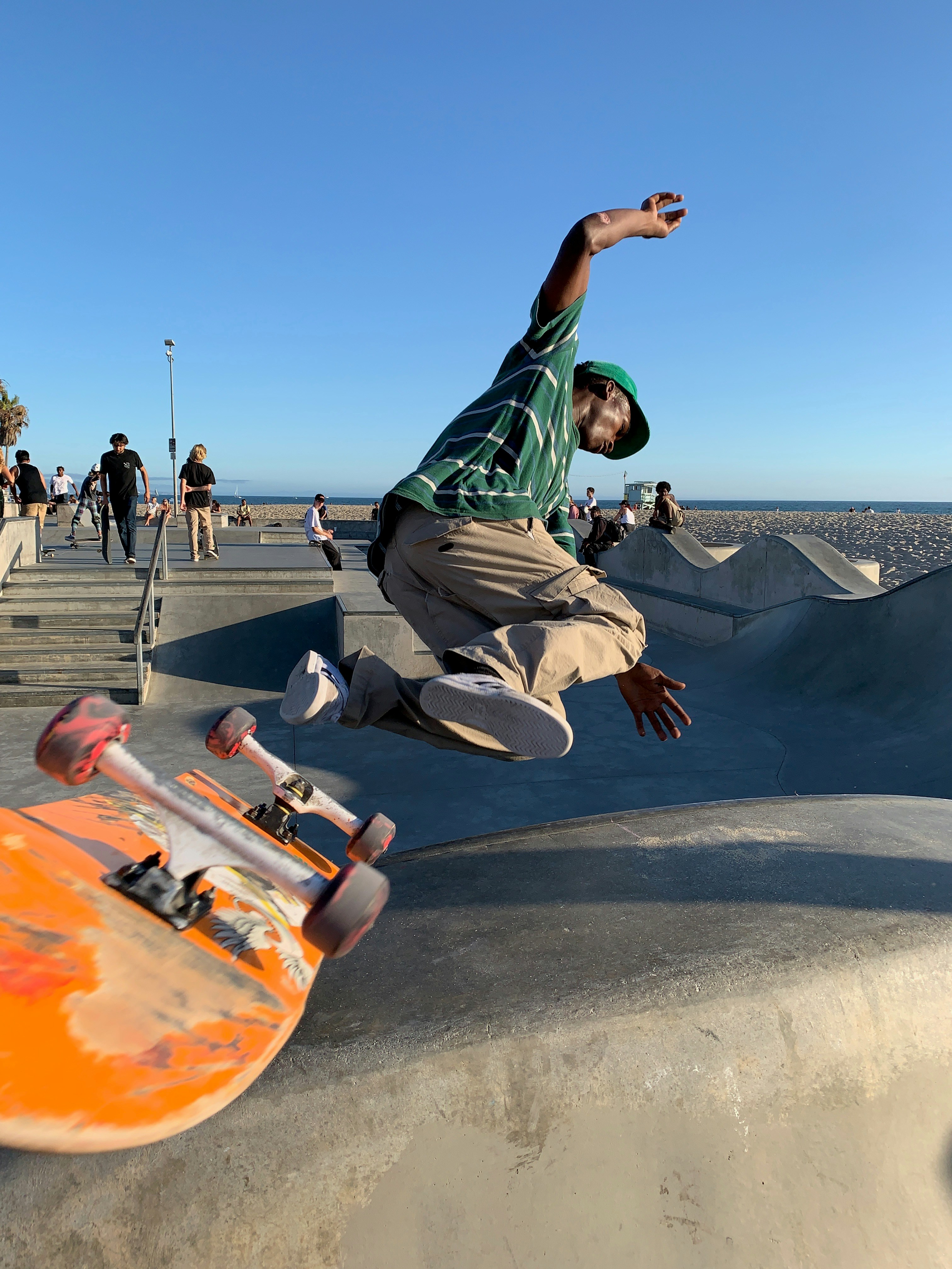 Skateboarder performing an aerial trick above a skate park with spectators in the background. The skateboard is prominently featured in the foreground.