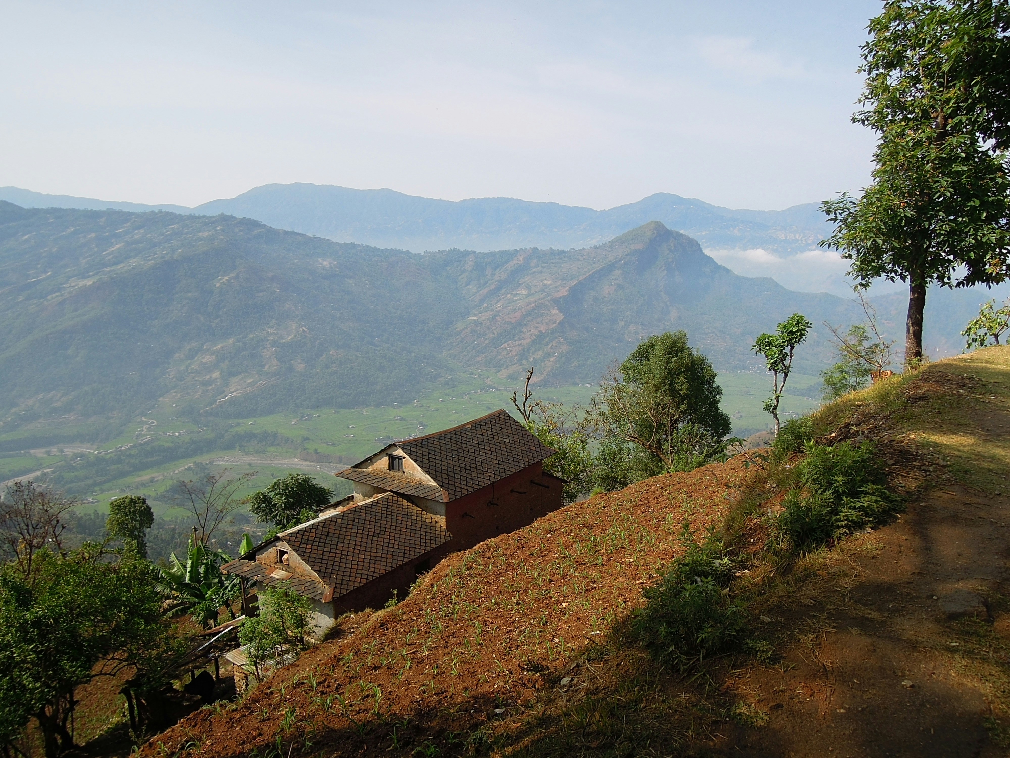 Photograph of a hillside village with terraced houses along a dusty slope, framed by trees and distant blue mountains.