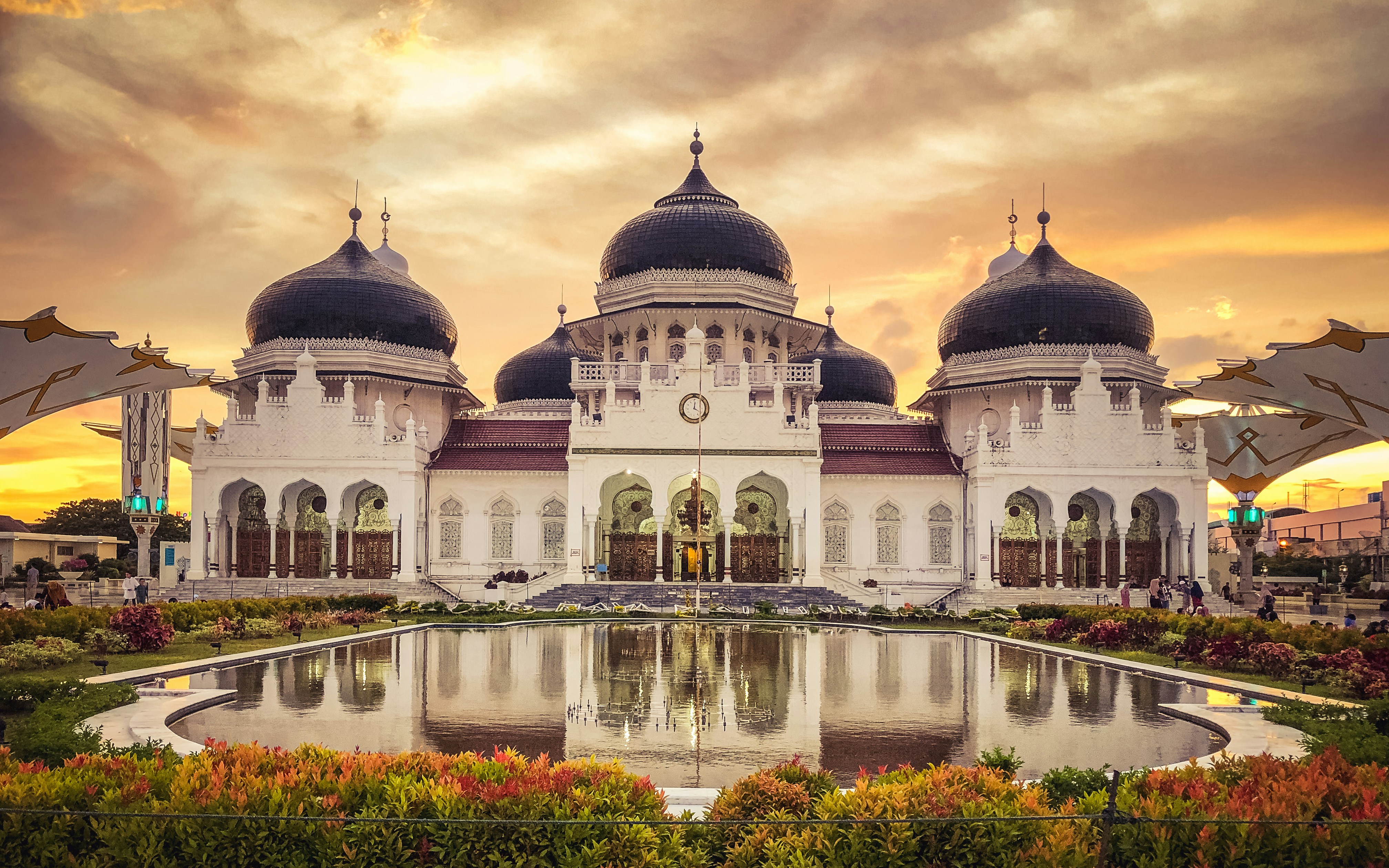 Majestic mosque reflecting in serene waters, framed by vibrant flower beds under a colorful sunset.