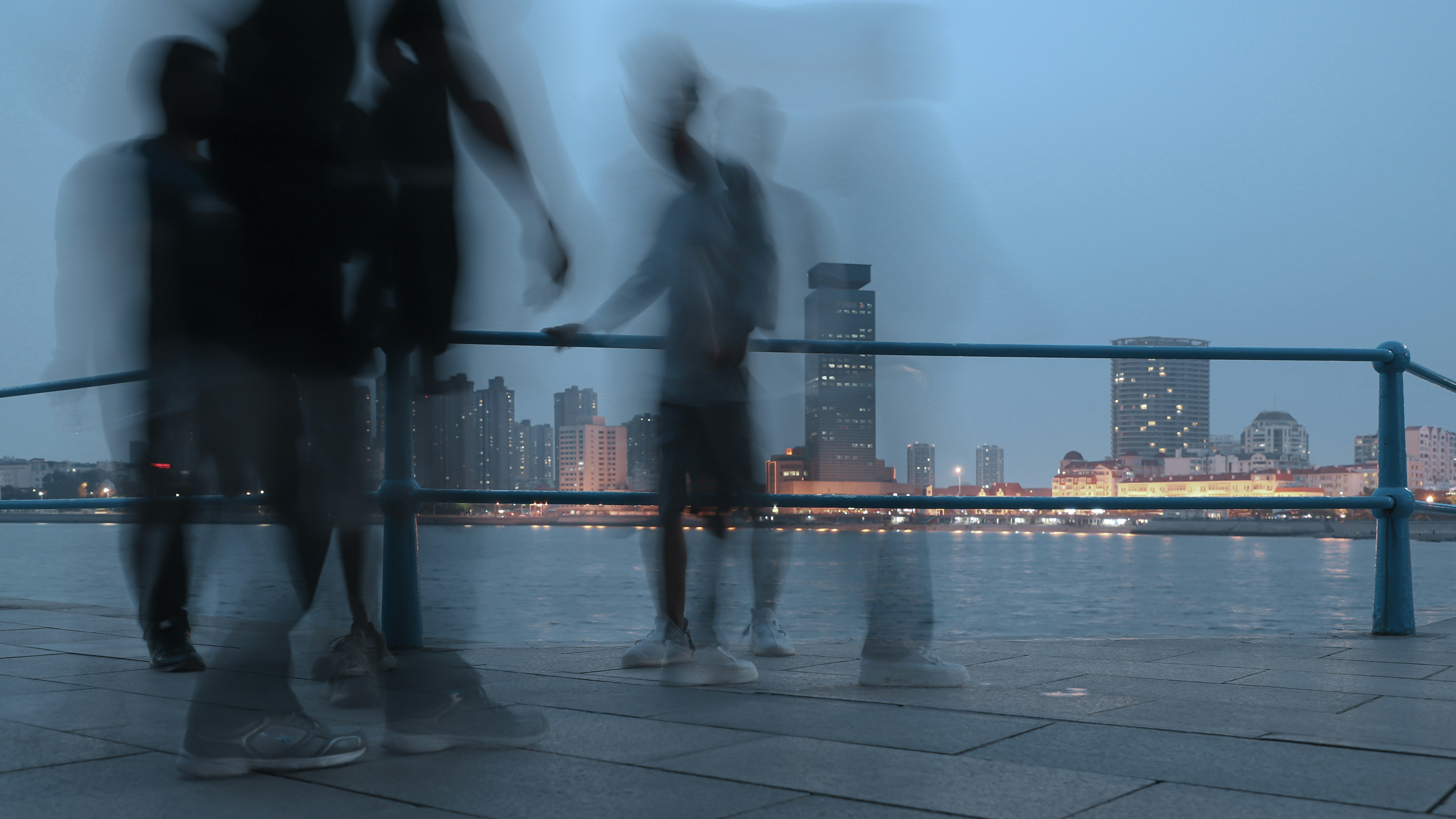 Blurred figures walking along a waterfront promenade with city skyline in the background at dusk.