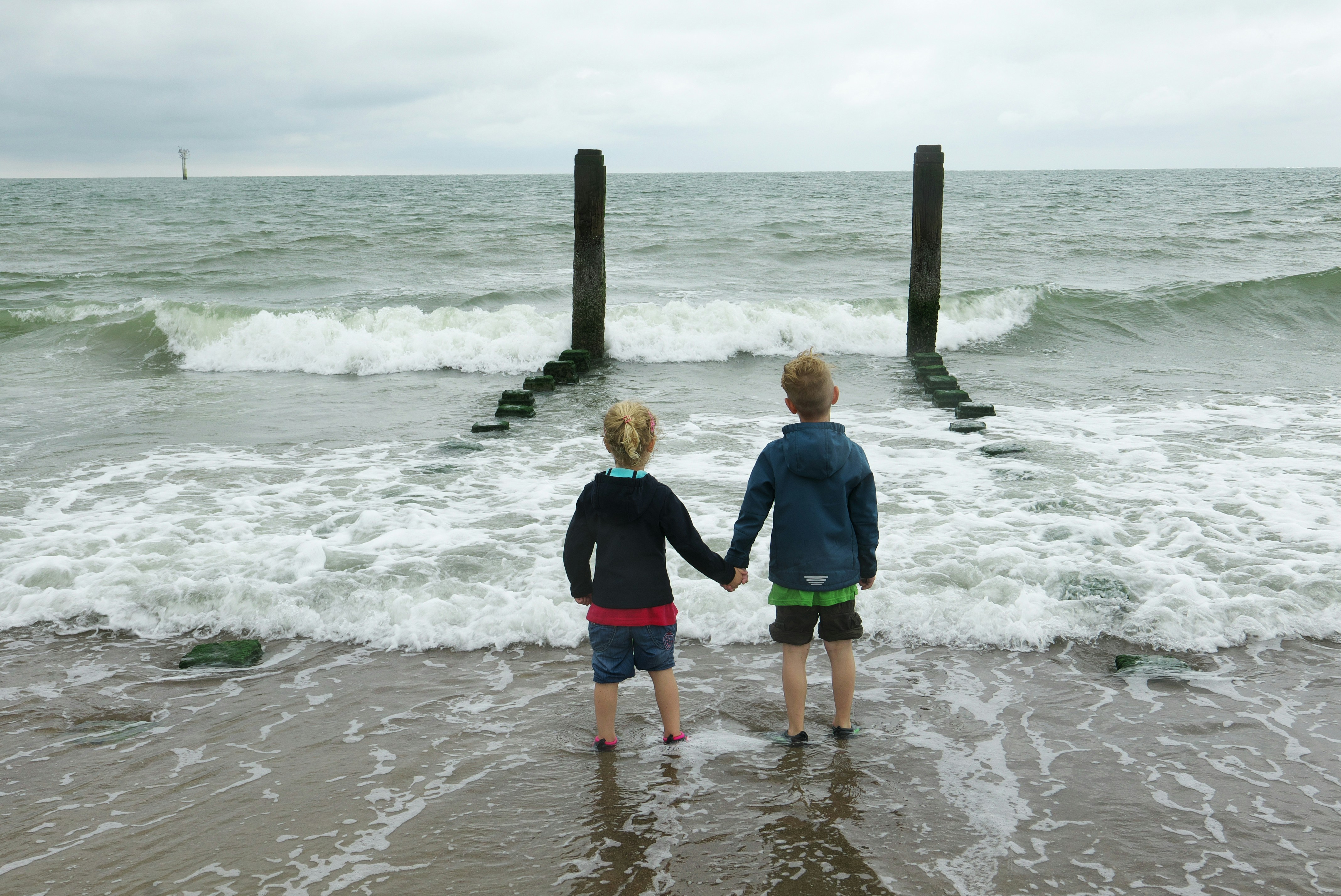 Two children holding hands at the water's edge, gazing at the waves and wooden posts emerging from the sea. The scene captures a moment of curiosity and adventure.