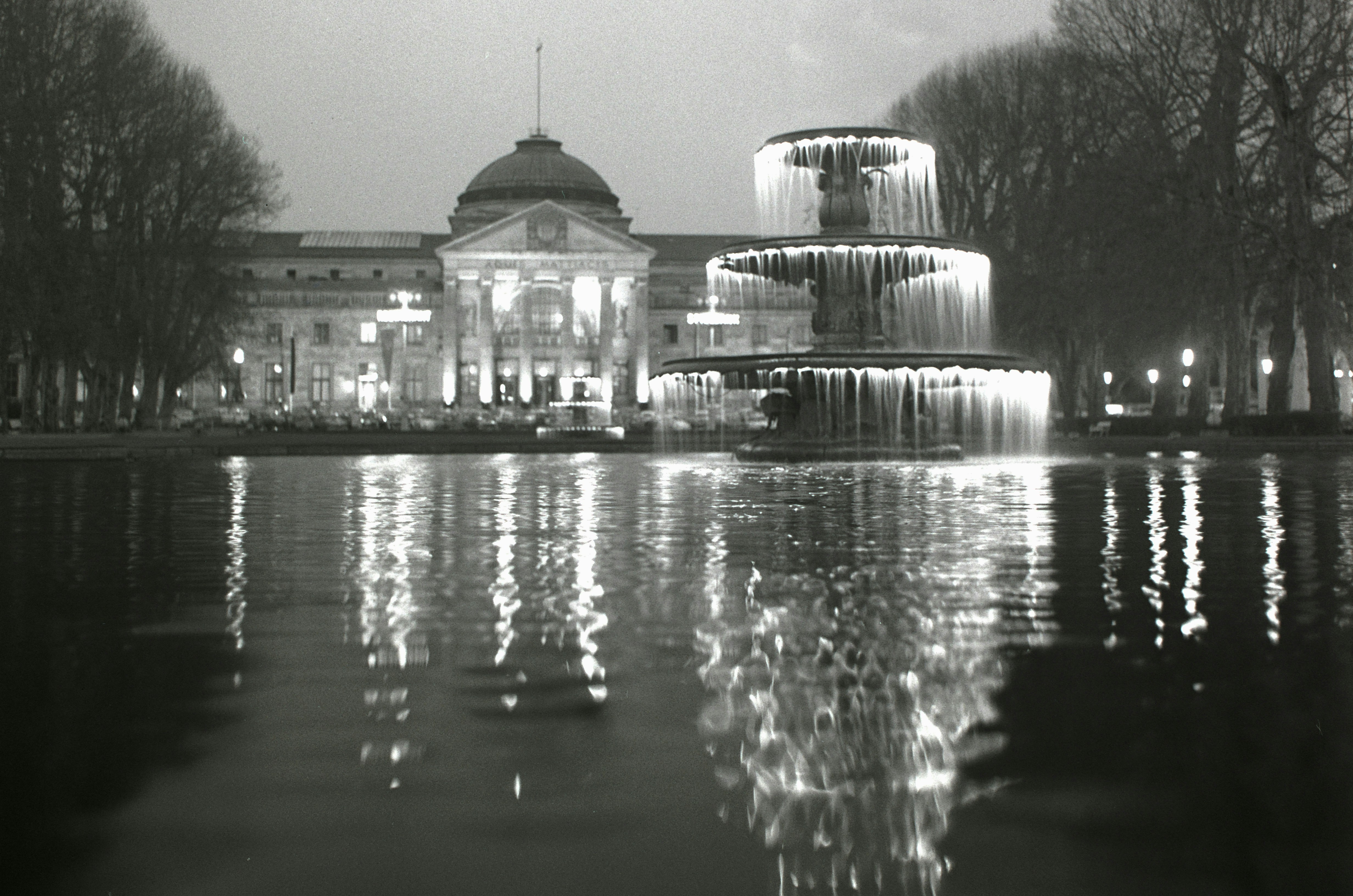 Grayscale image of a grand building with a dome, reflected in a tranquil body of water at night.