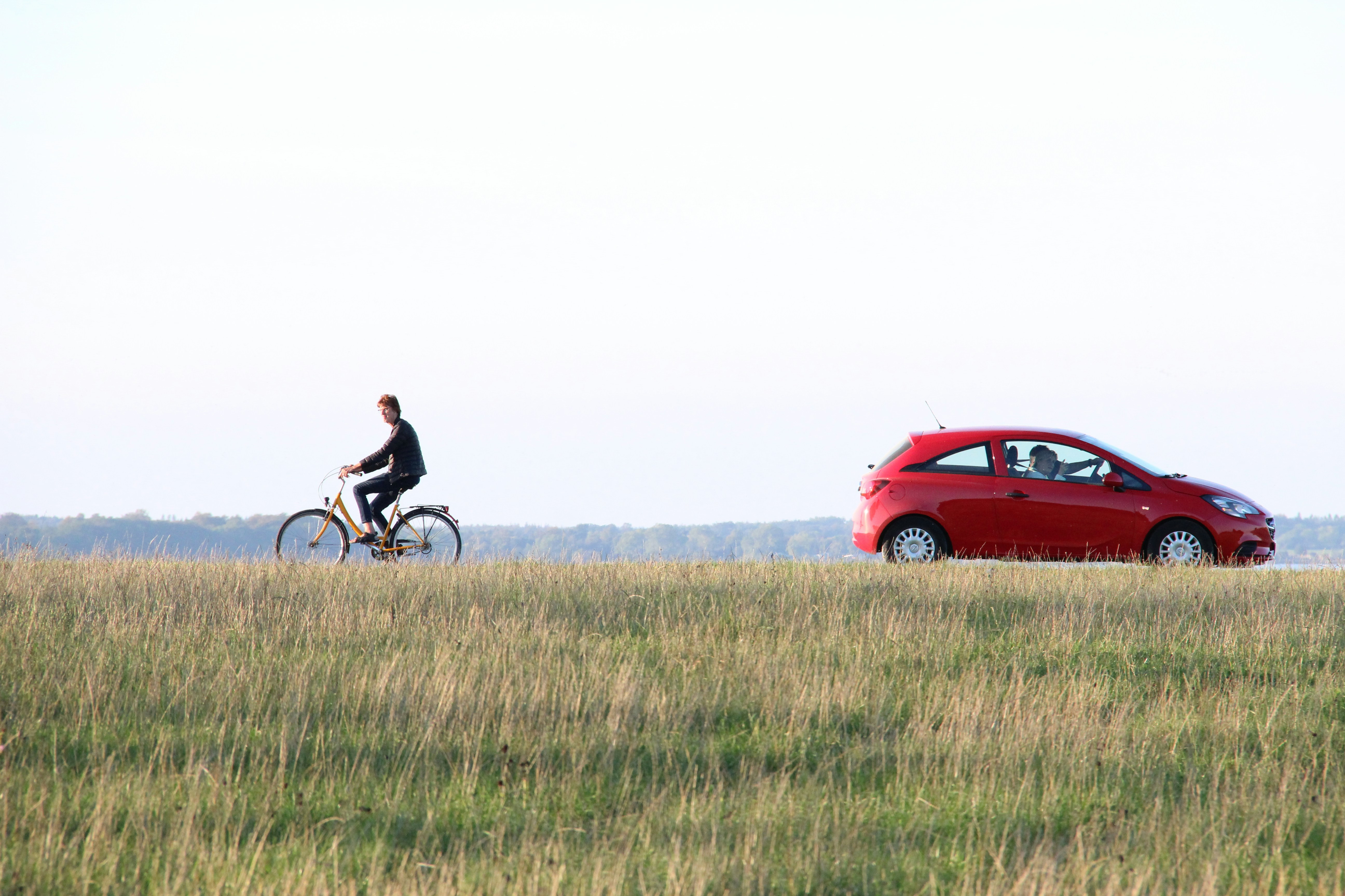 A cyclist rides along a grassy expanse beside a red car, showcasing the contrast between human-powered and motorized transport. The scene captures a serene moment of travel.