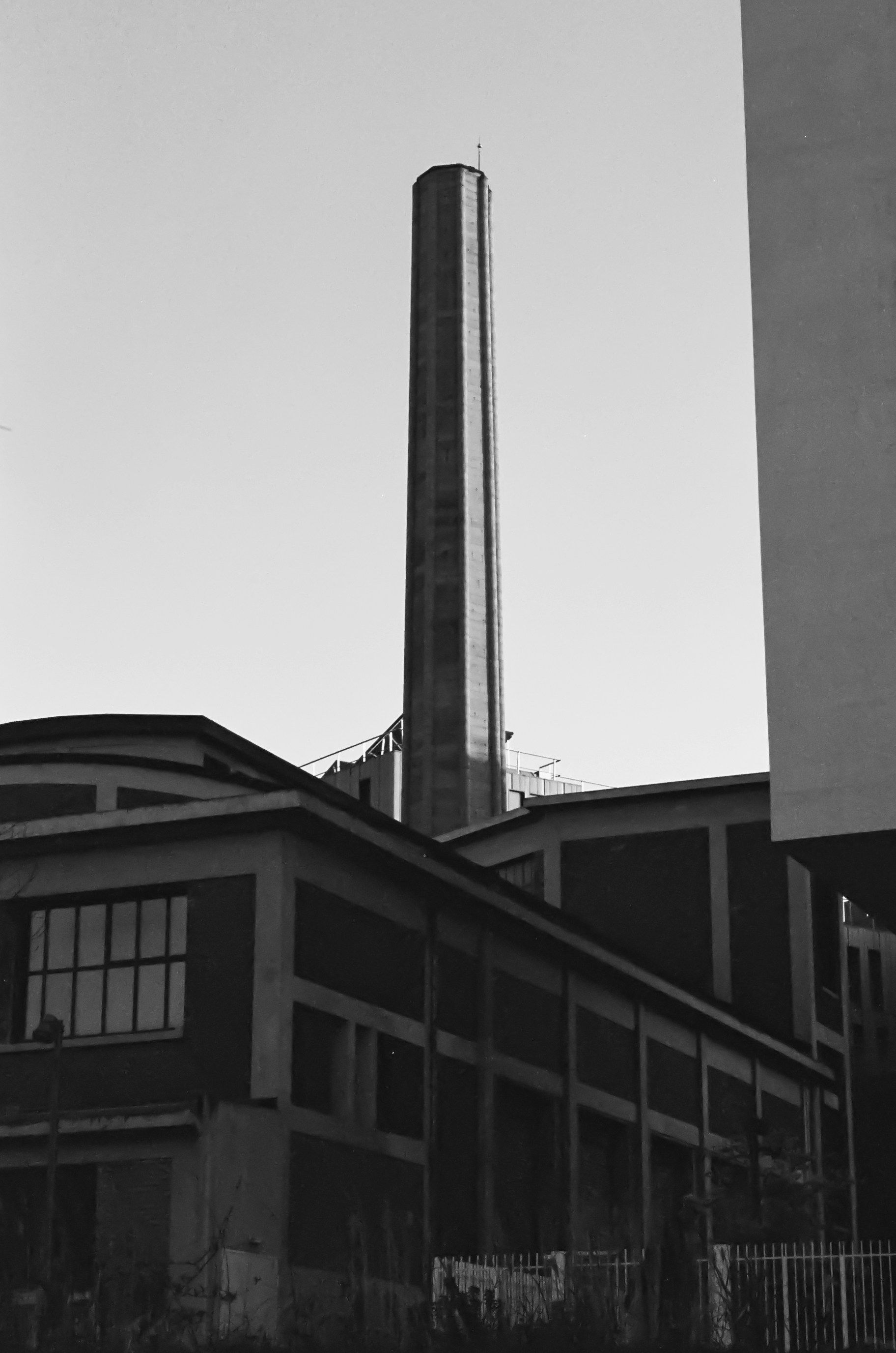 Black-and-white photograph of a factory complex dominated by a tall chimney against a pale sky.