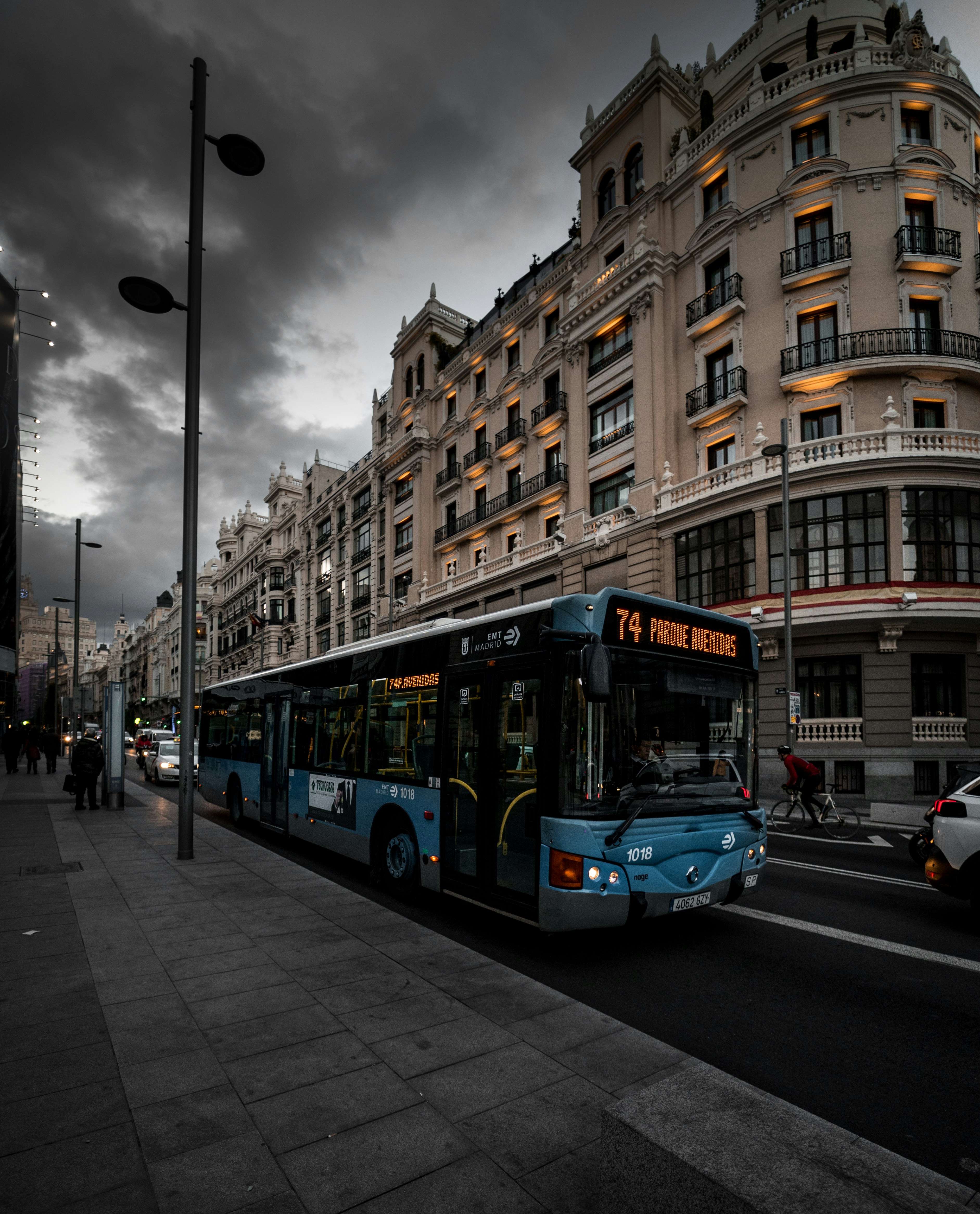A city bus navigating a bustling street under a dramatic sky, flanked by elegant architecture. The scene captures the essence of urban life in motion.