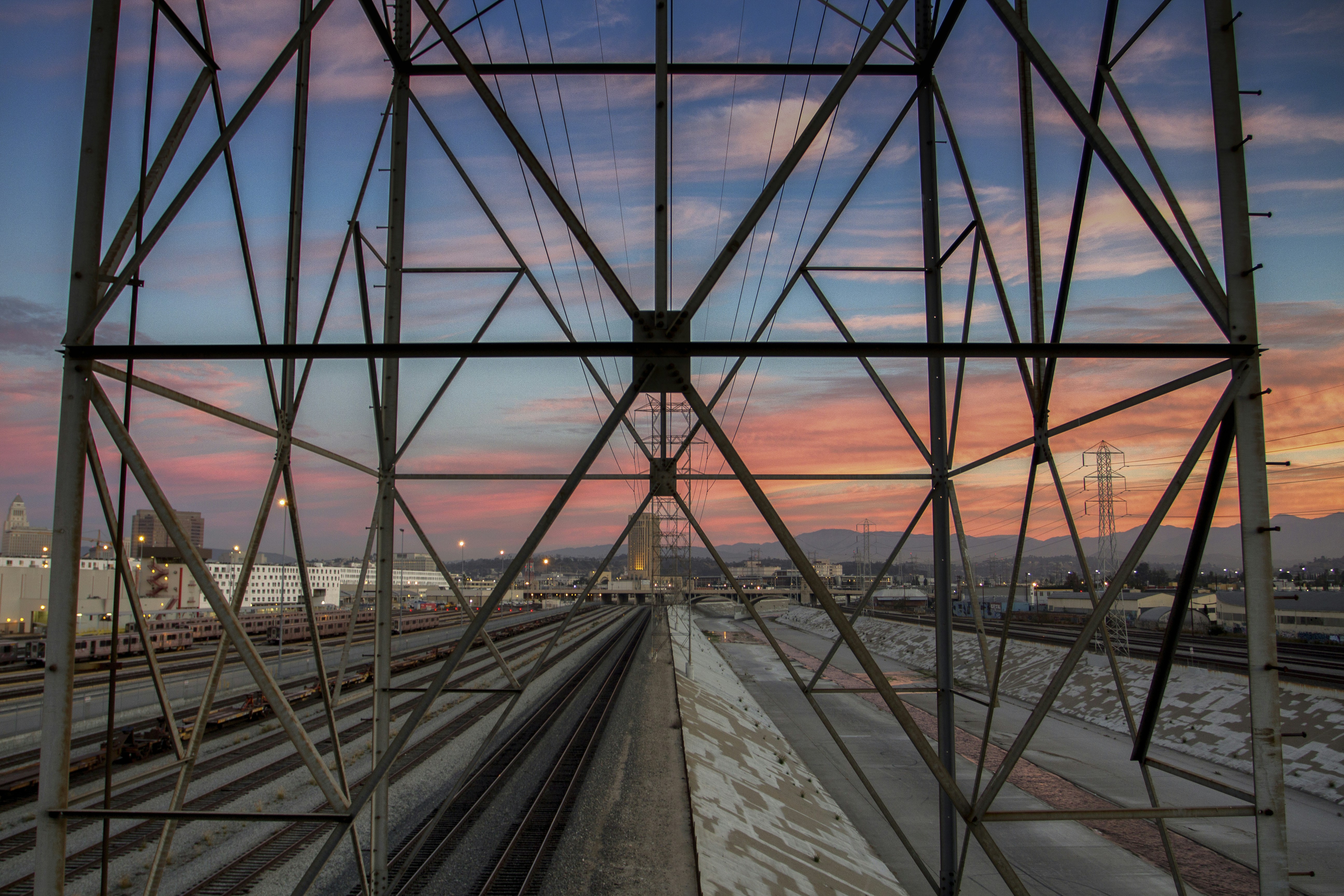 Steel lattice tower framing a vibrant sunset over railway tracks and distant mountains.