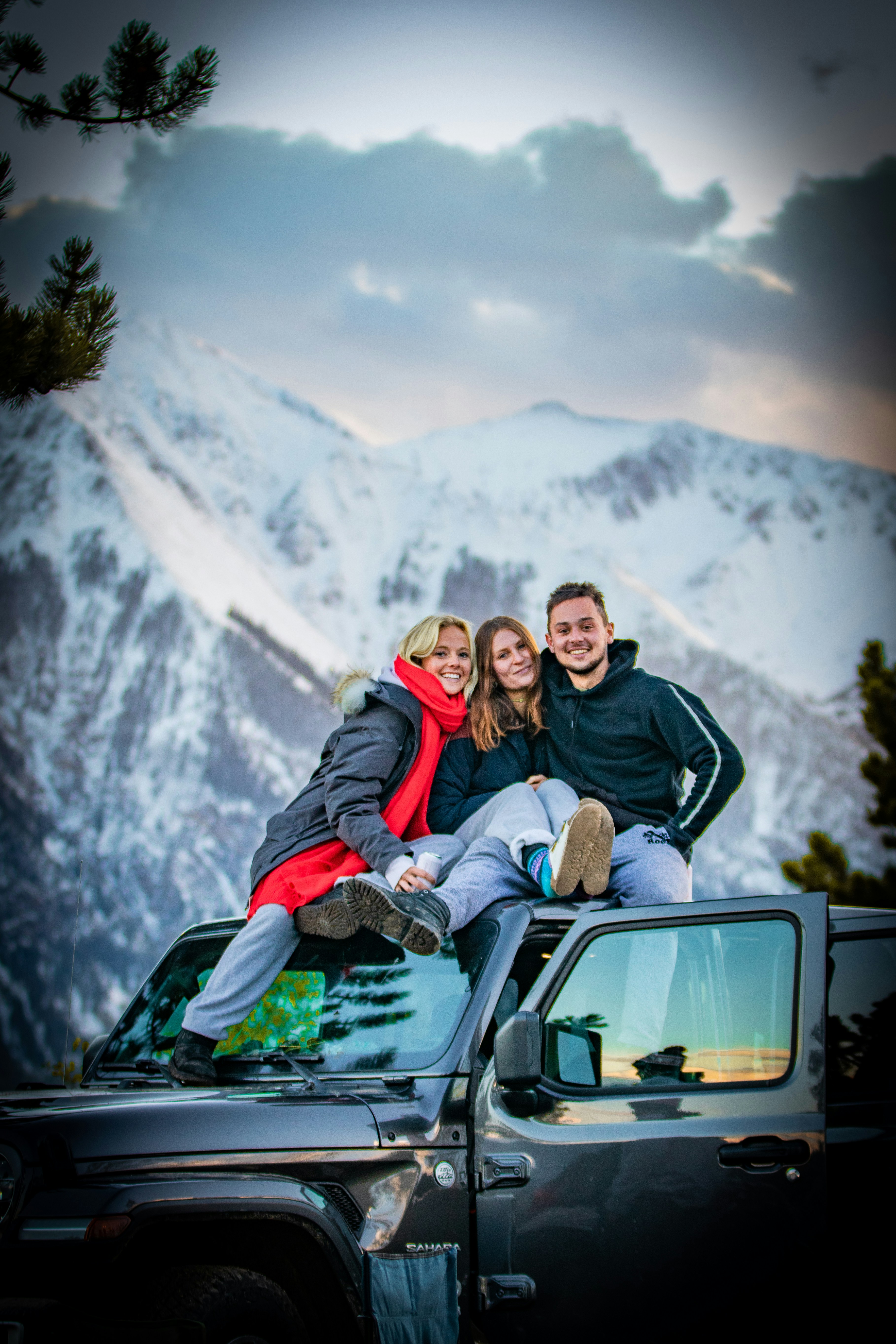 Three Friends Perched on a Rugged SUV Against a Snowy Alpine Backdrop