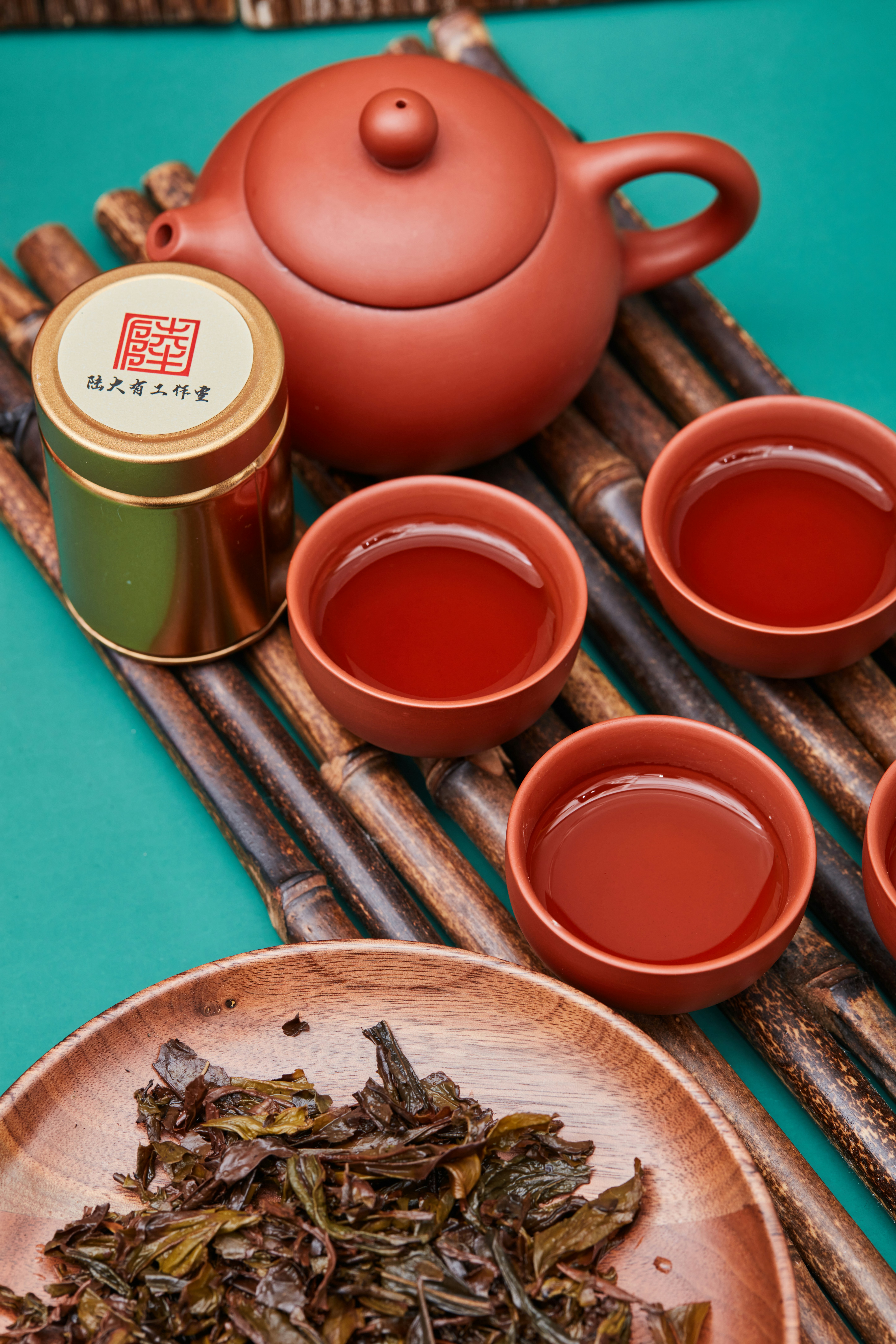 Teapot and cups arranged on bamboo mat with dried tea leaves on wooden plate, showcasing a traditional tea ceremony setup.