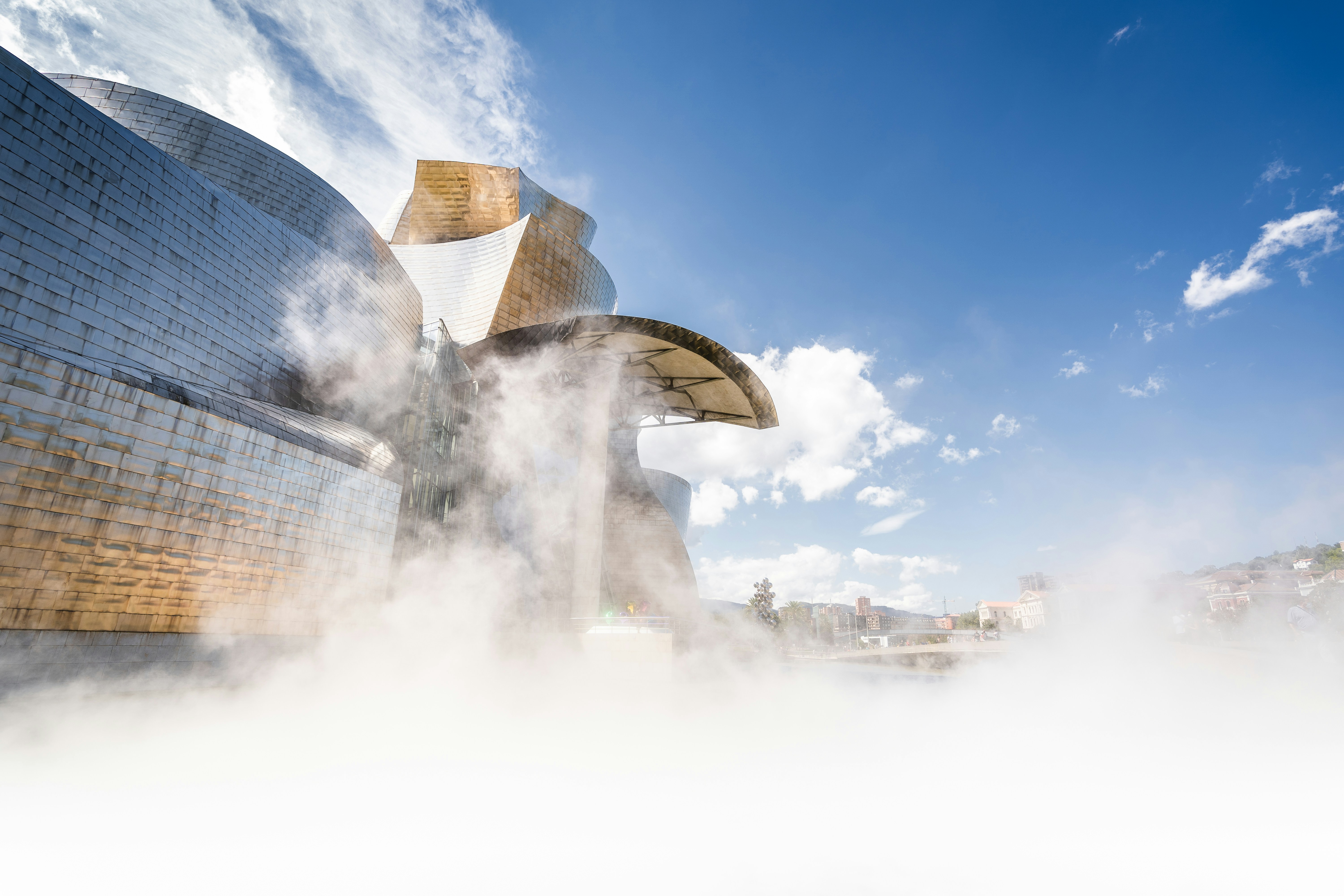 Guggenheim Museum Bilbao's metallic curves enveloped by ethereal fog beneath a bright blue sky.