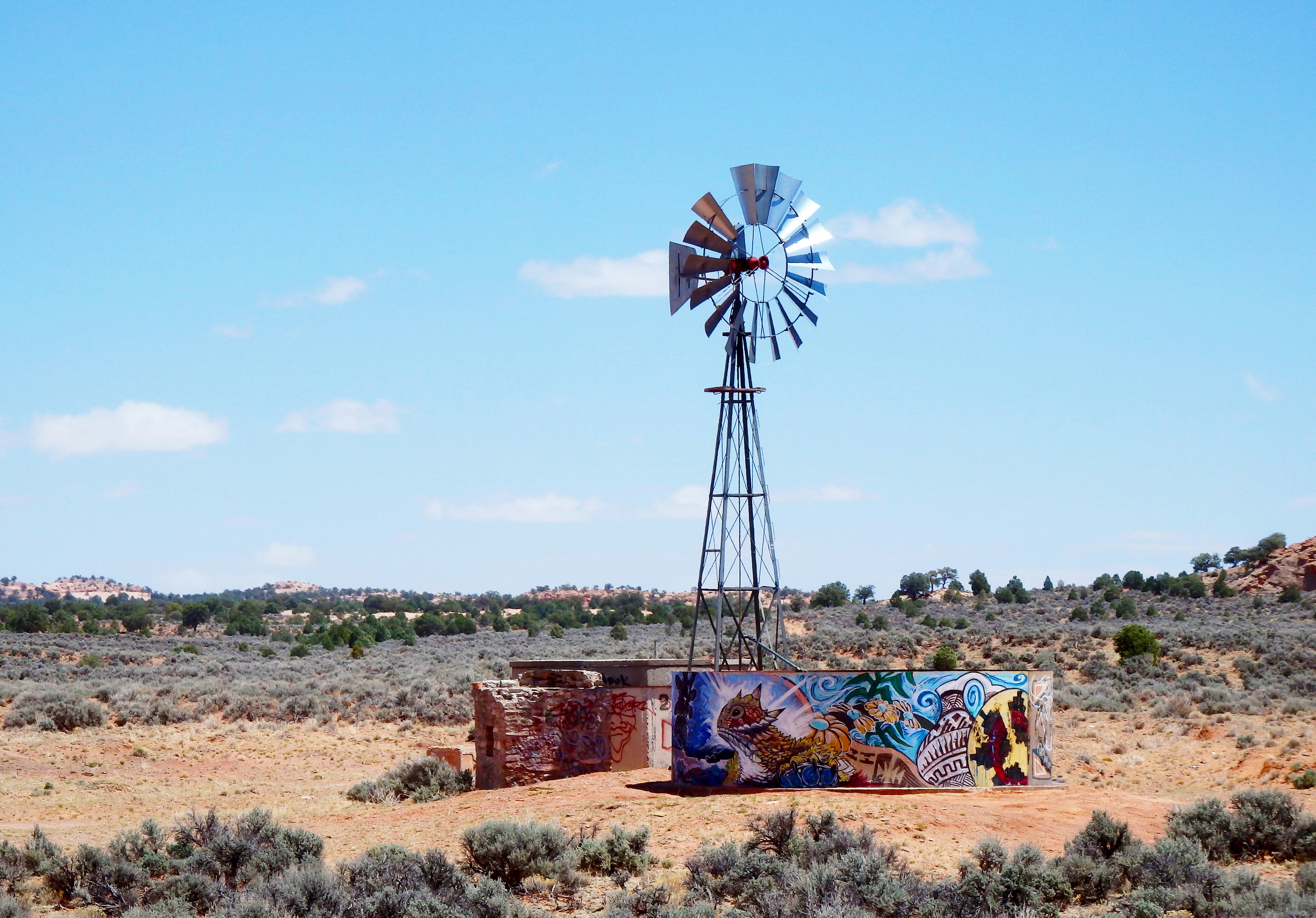 Windmill rises behind a graffiti-covered concrete shelter in a sparse desert scene beneath a bright blue sky.
