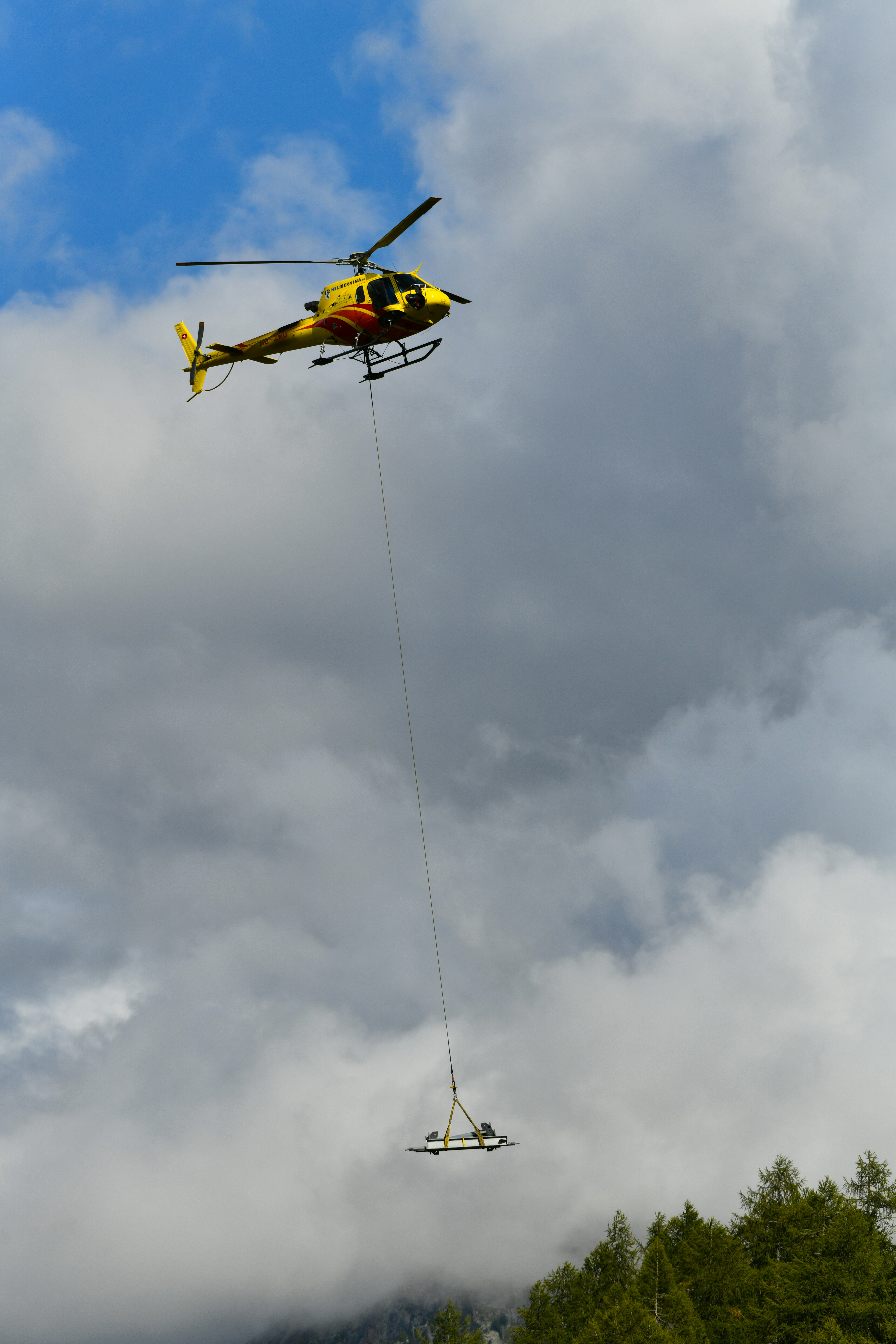 Yellow rescue helicopter hoisting unpowered gondola against dramatic clouds