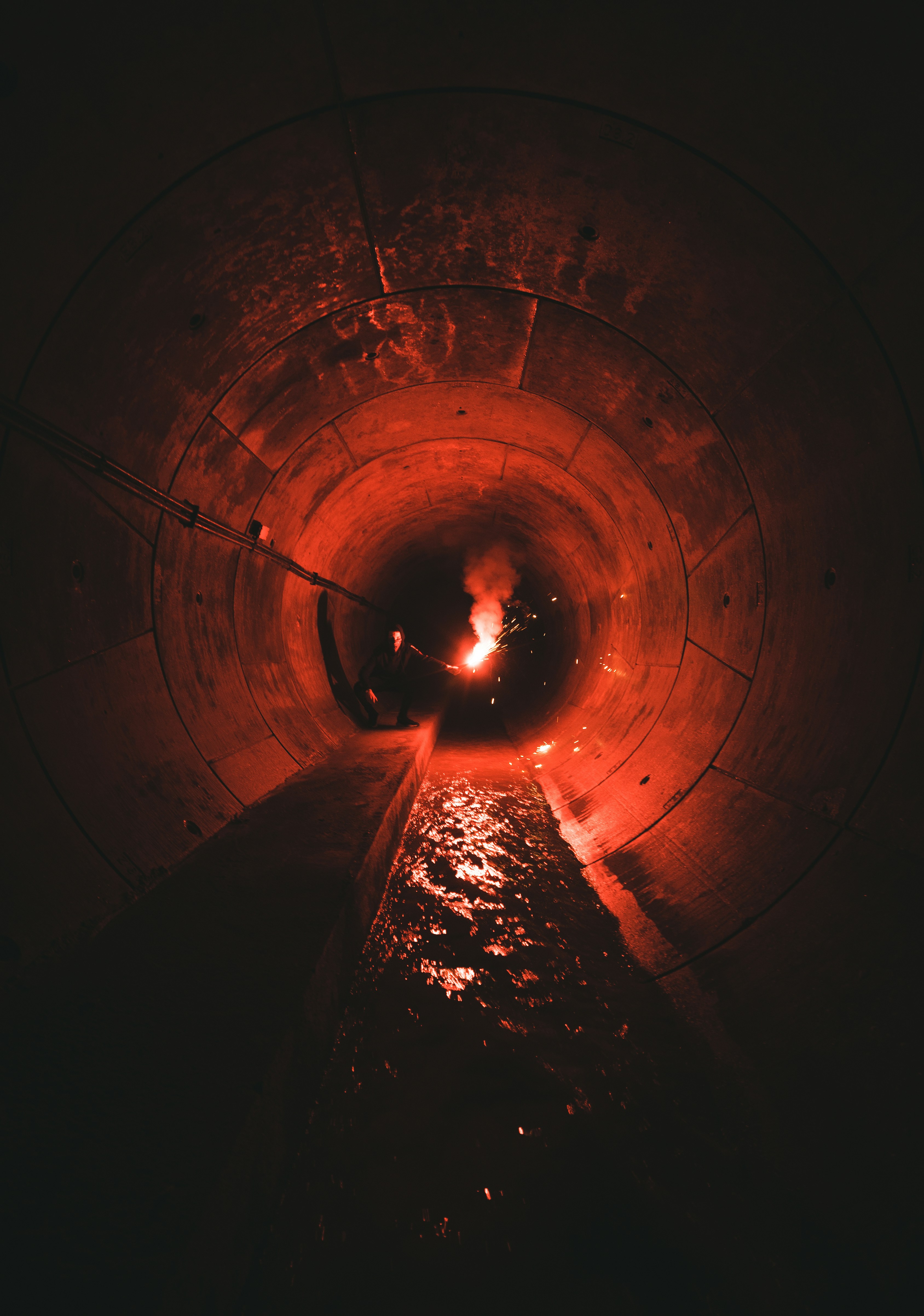 A figure ignites a flare inside a dark, circular tunnel, casting an eerie red glow on the concrete walls. The water reflects the light, enhancing the sense of depth.