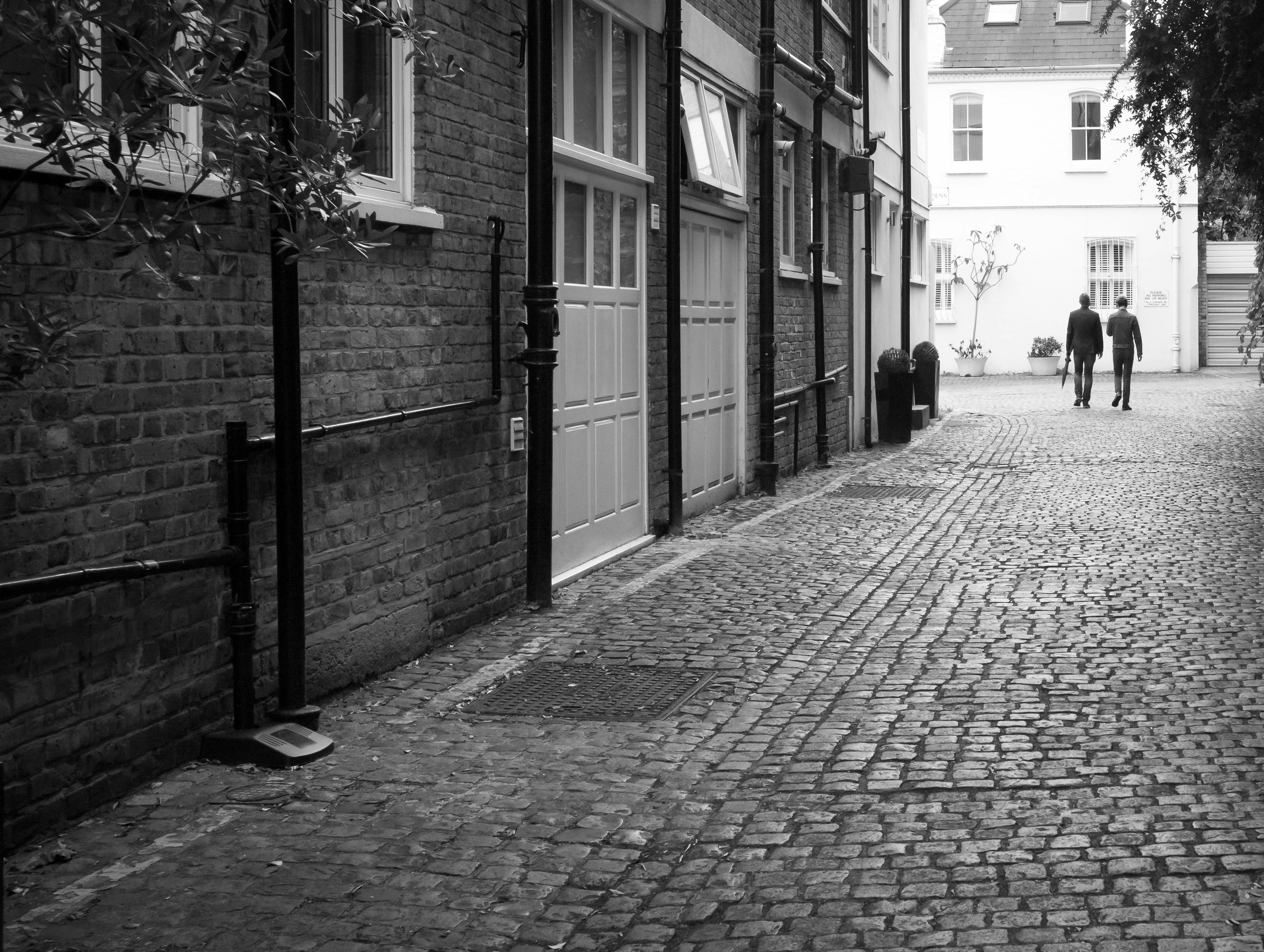 Cobblestone alleyway framed by brick buildings, with two figures walking in the distance. The scene exudes a sense of quiet introspection.