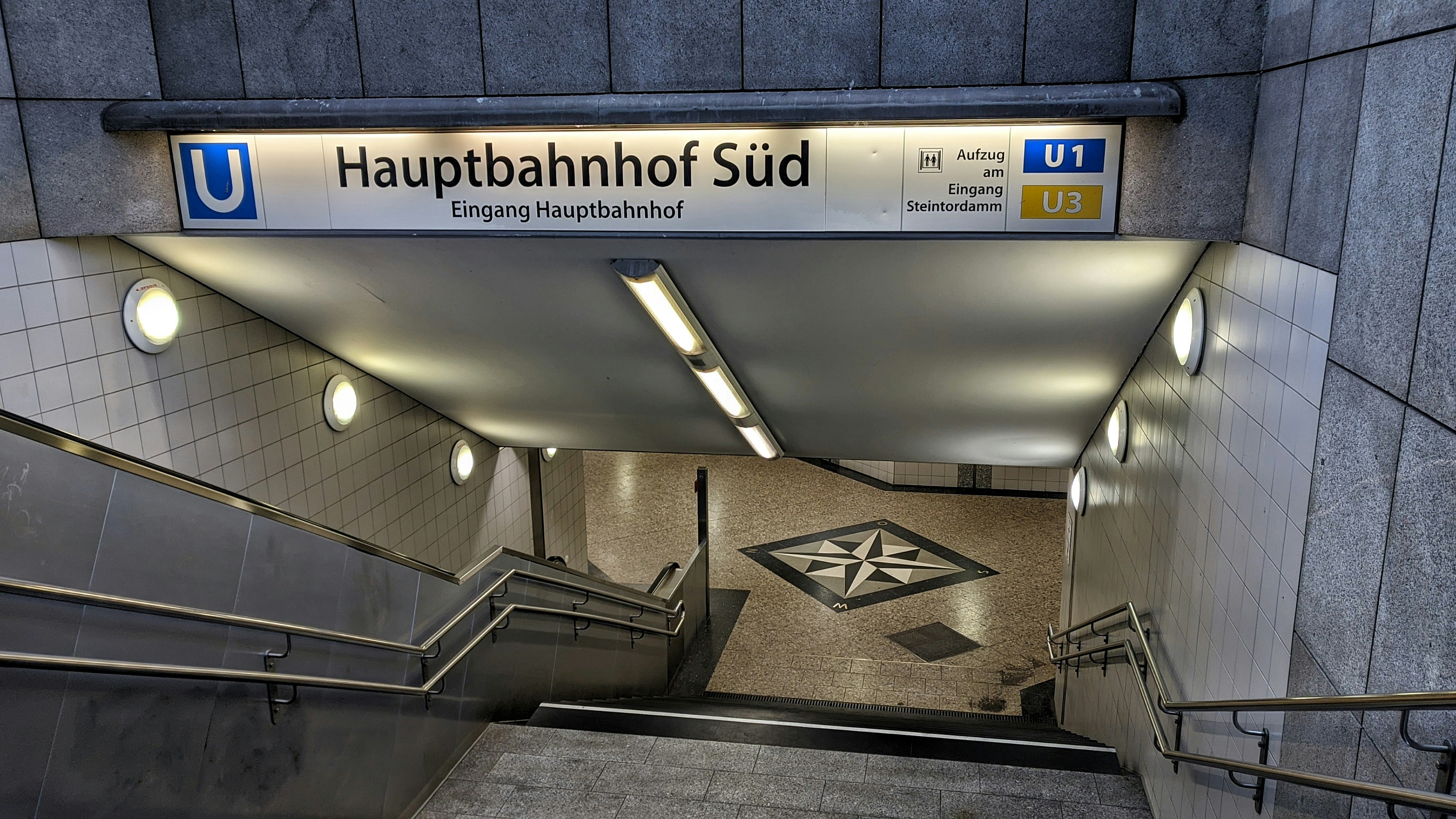 Staircase leading down to the Hauptbahnhof Süd subway station, featuring a prominent sign and geometric floor design.