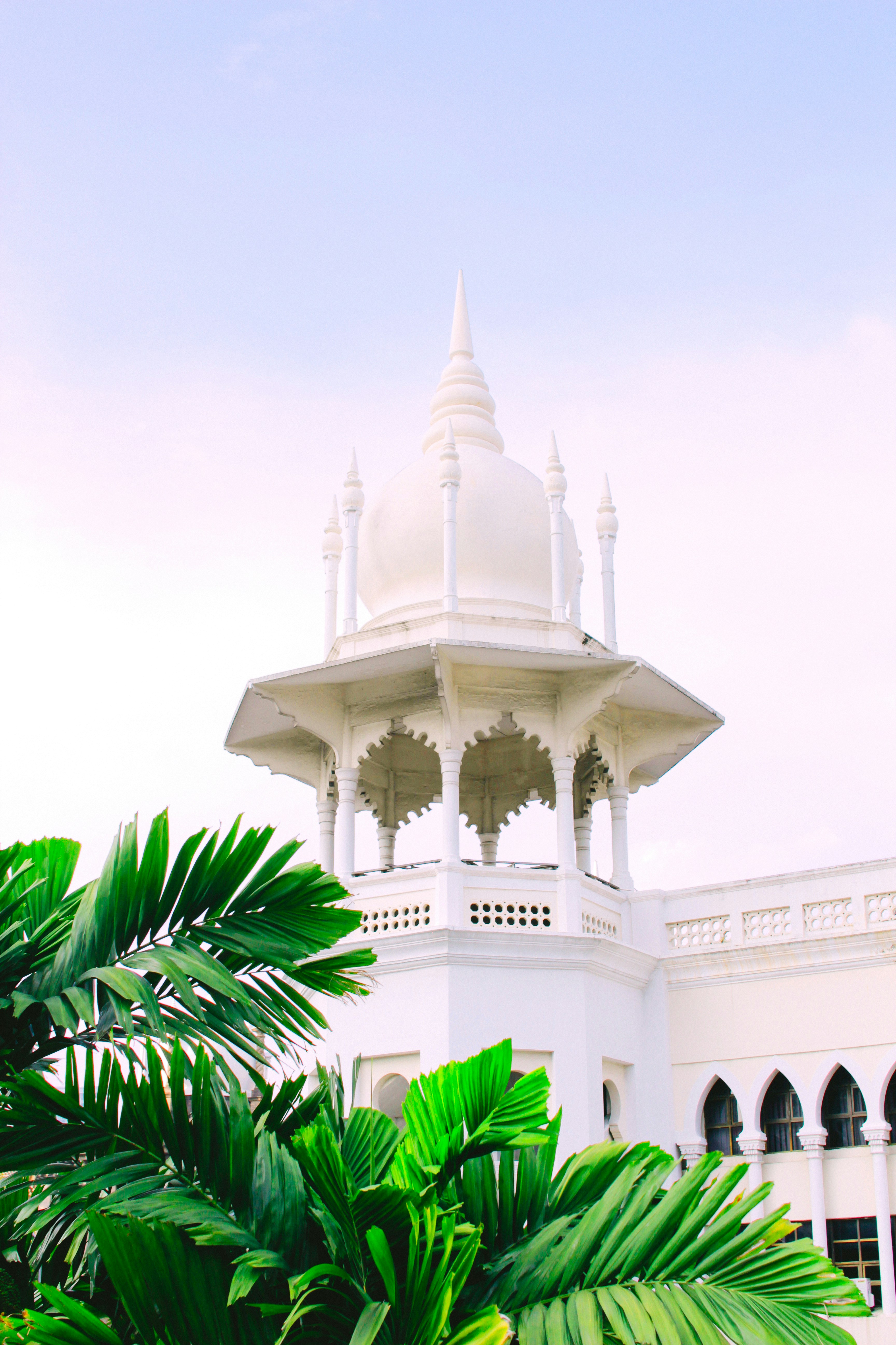 Elegant white pavilion adorned with intricate arches and a domed roof, framed by lush green foliage under a soft blue sky.