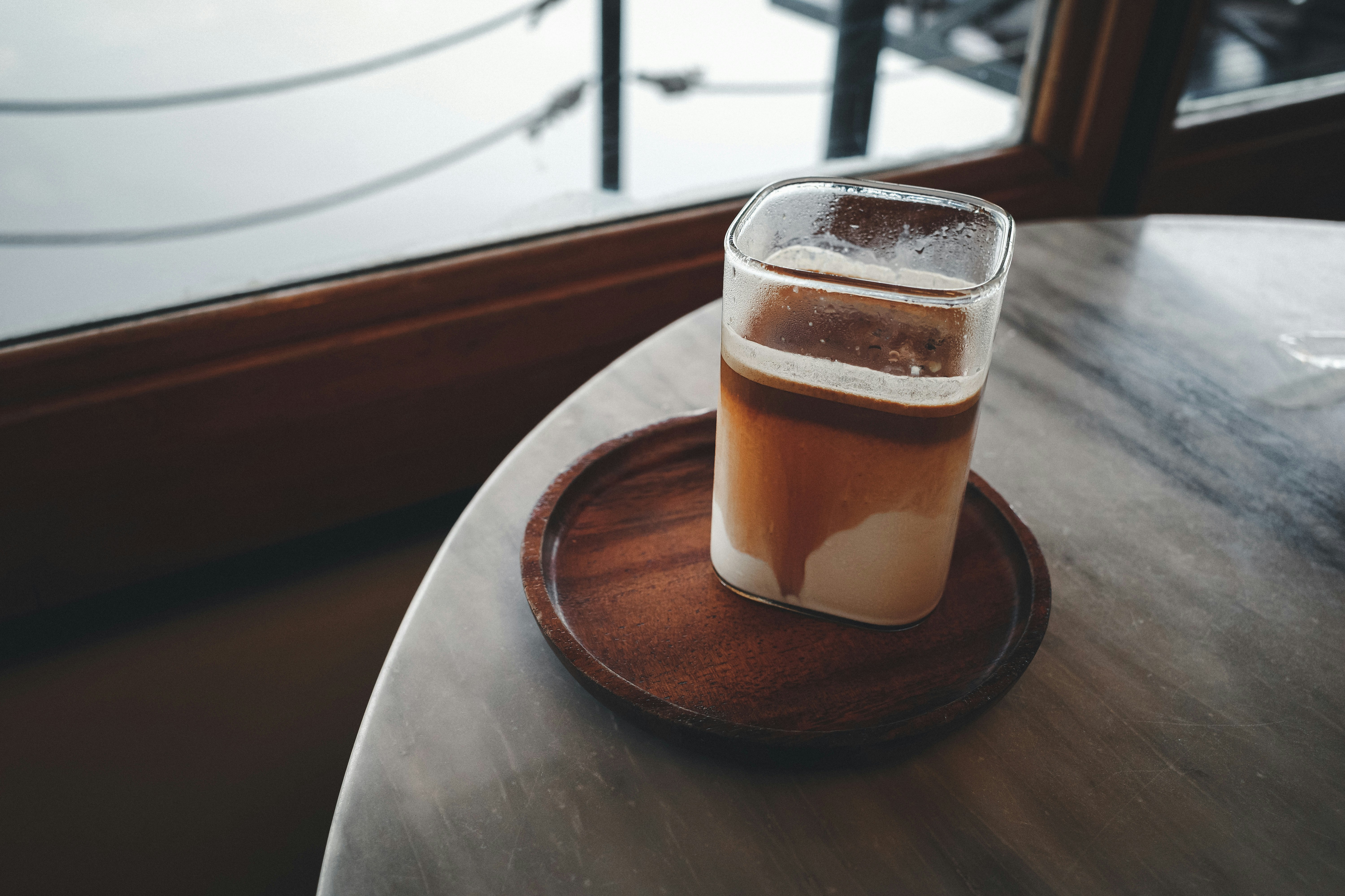 A glass of iced coffee rests on a wooden coaster, with reflections of a serene outdoor scene visible through the window. The creamy layers of the drink create an inviting visual contrast.
