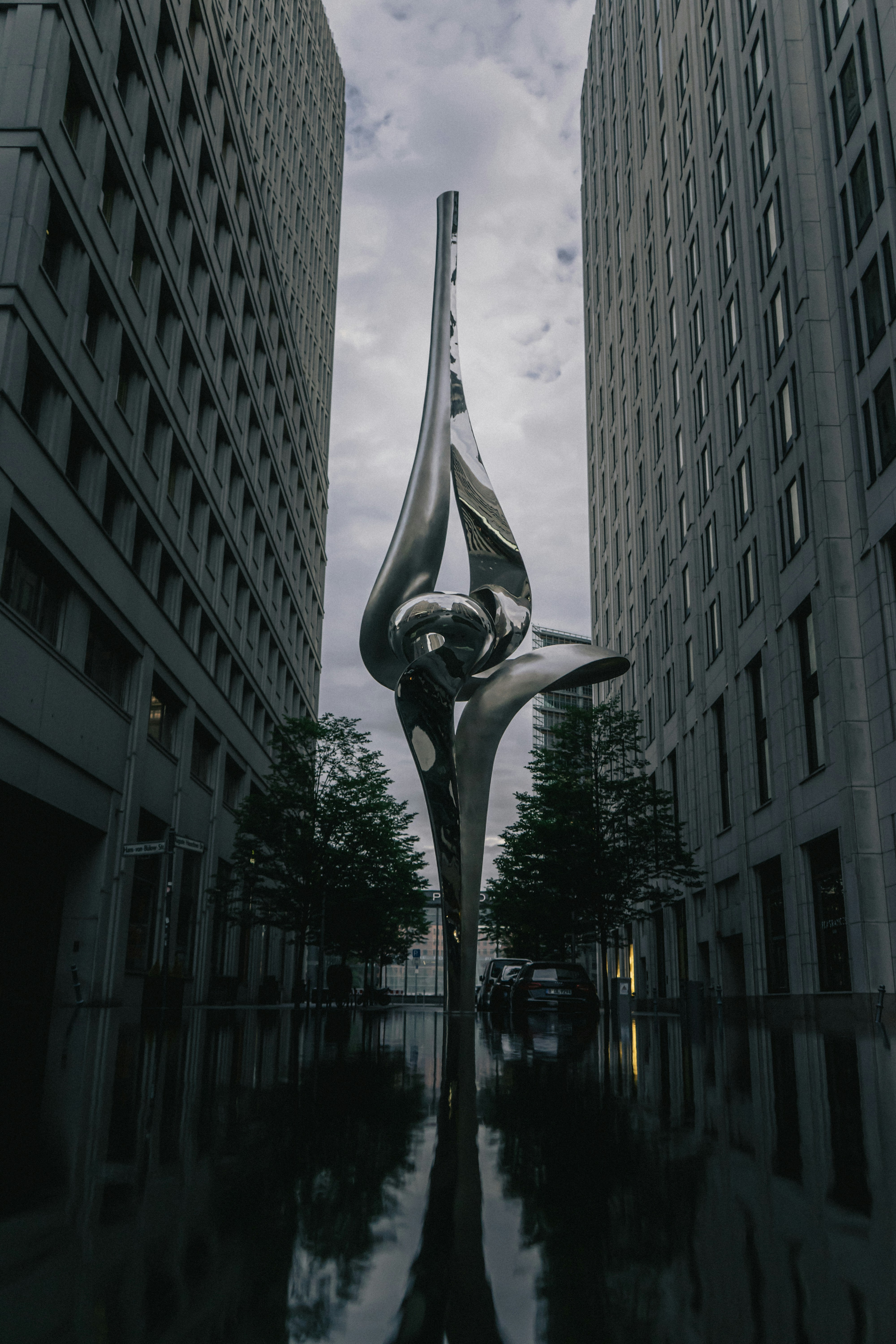 A sleek metallic sculpture stands tall between two modern buildings, reflecting the cloudy sky and surrounding greenery.