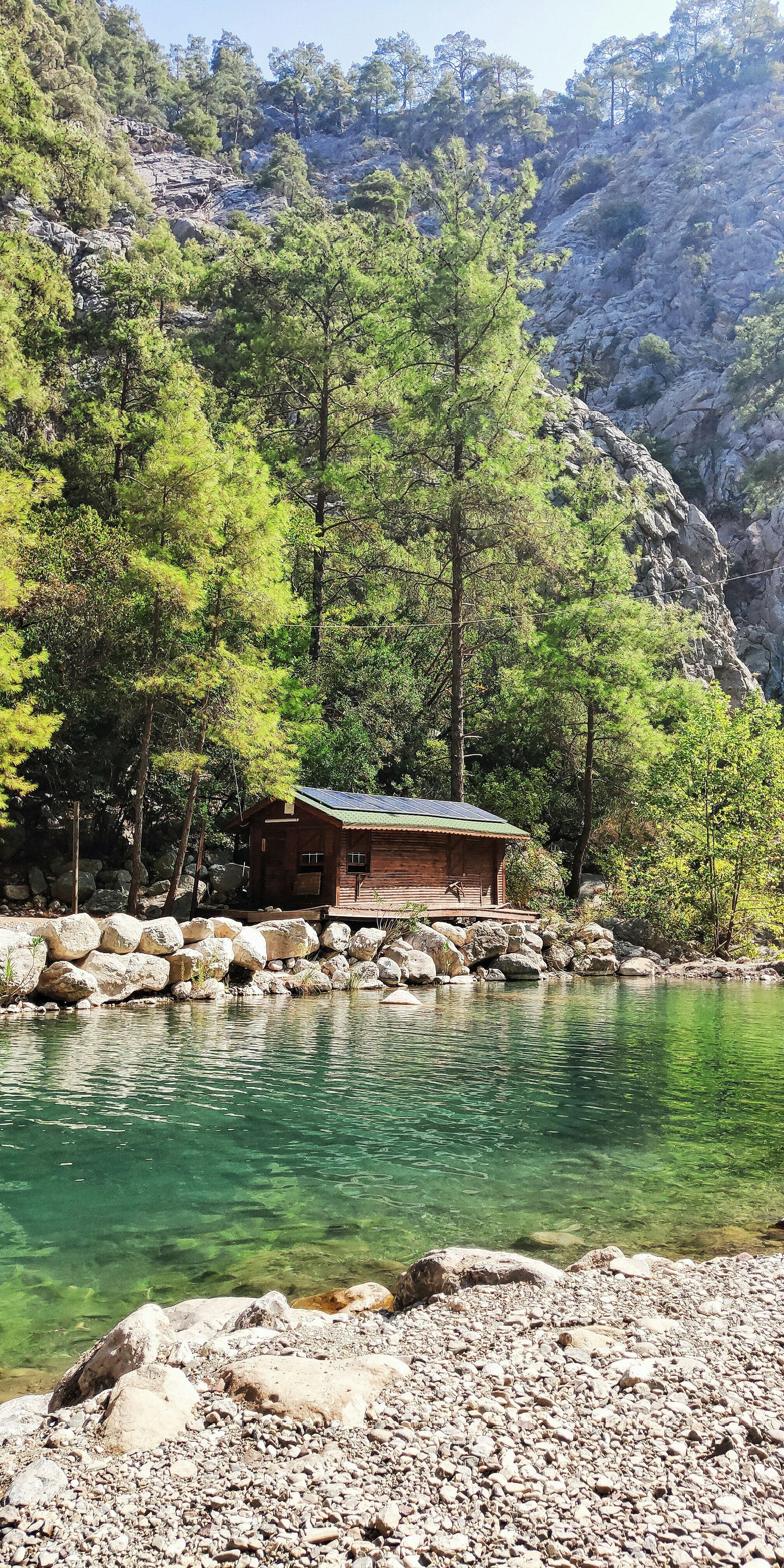 Wooden cabin nestled among trees beside a tranquil green river, surrounded by rocky terrain.