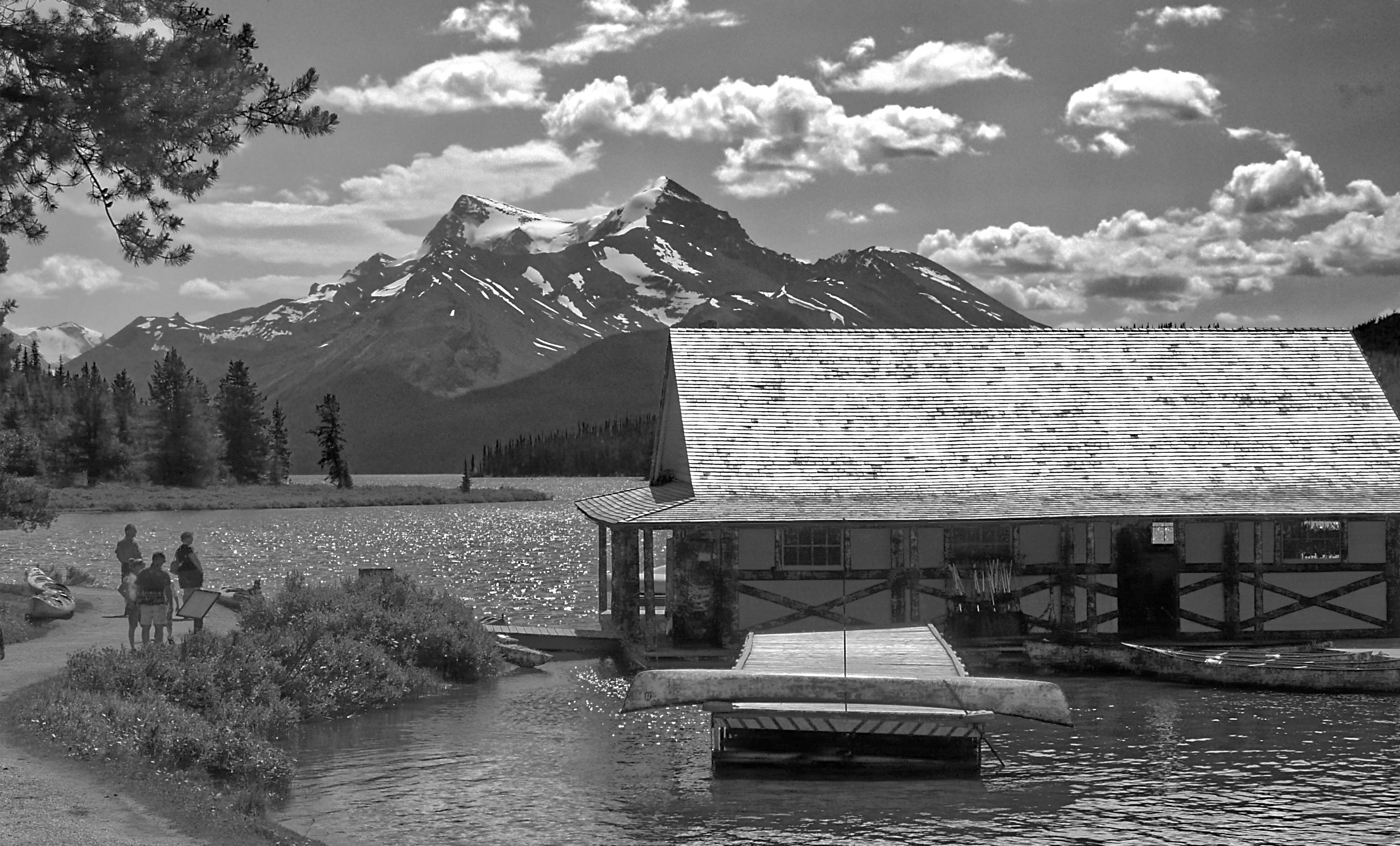 Snow-capped mountains rise behind a reflective lake with a rustic boathouse and dock; a small group sits near the shoreline, creating a calm, pastoral landscape.