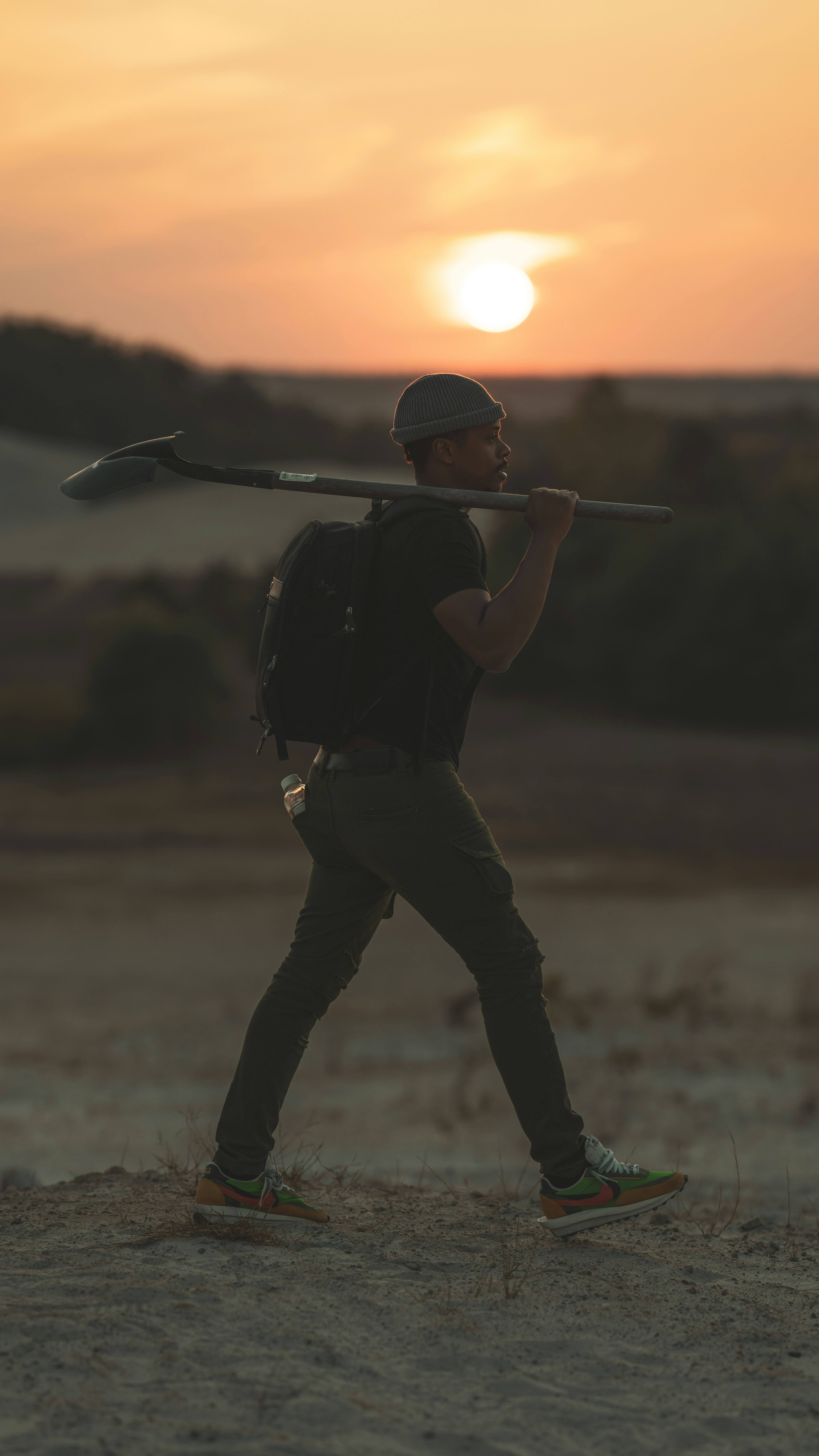 A determined individual strides across a sandy landscape, silhouetted against a glowing sunset, carrying a shovel. The scene captures the essence of exploration and purpose.