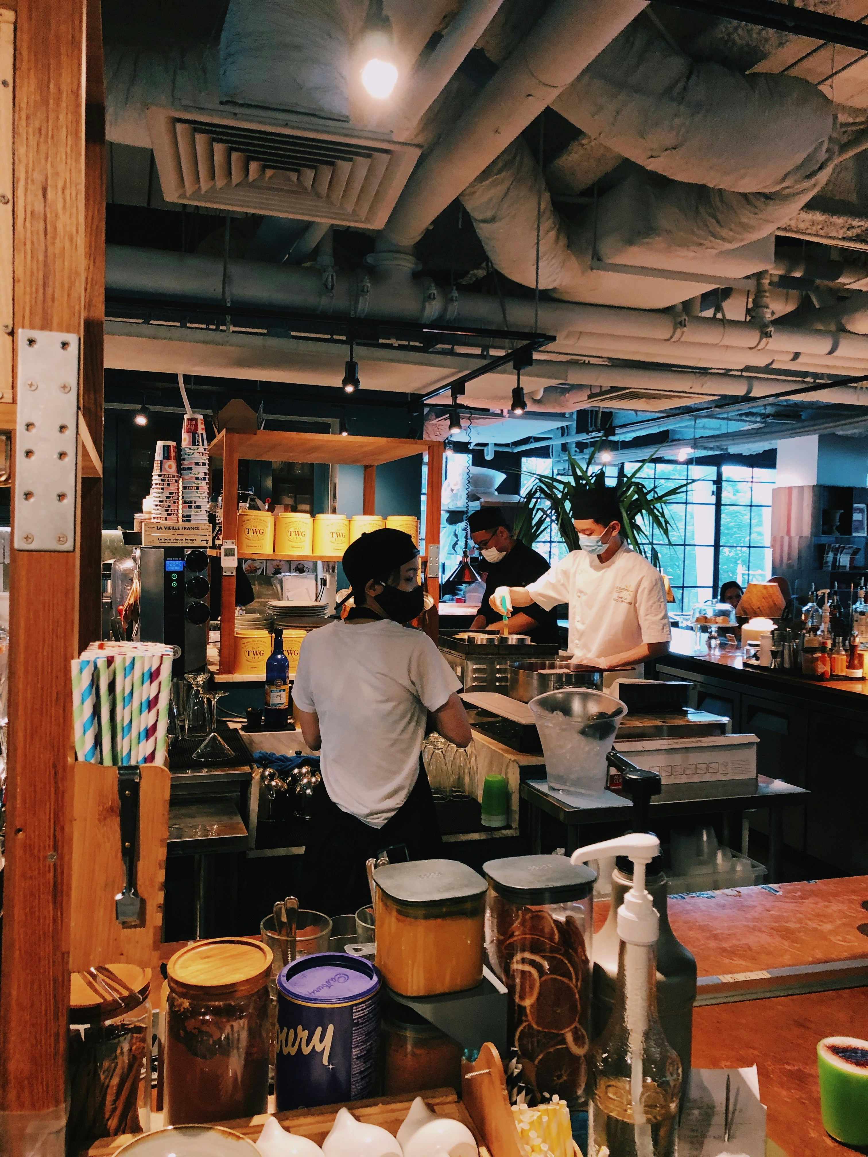Masked Baristas Orchestrate a Busy Café Kitchen Under Warm Industrial Lights