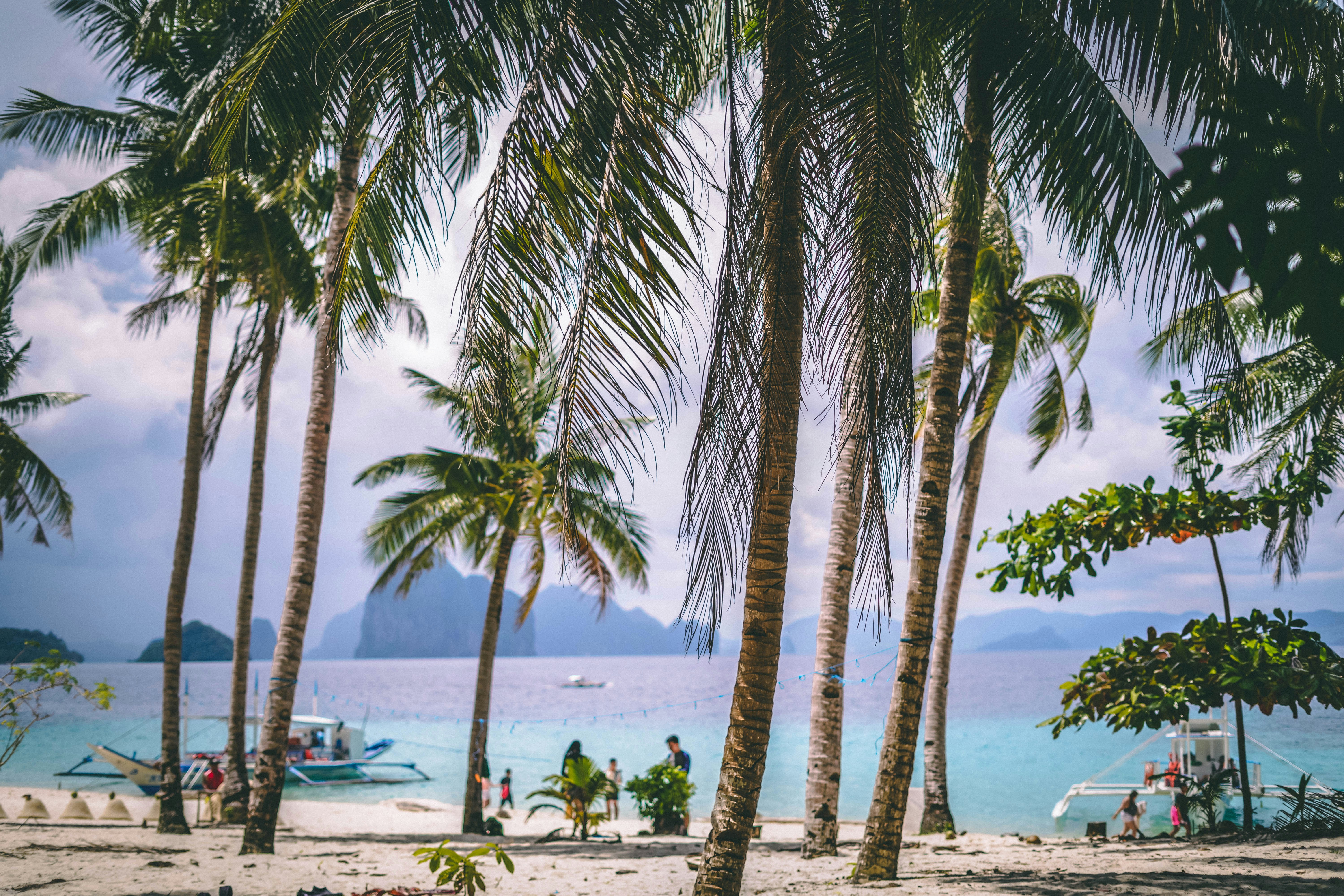 Palm trees frame a serene beach with boats on turquoise waters under a cloudy sky.
