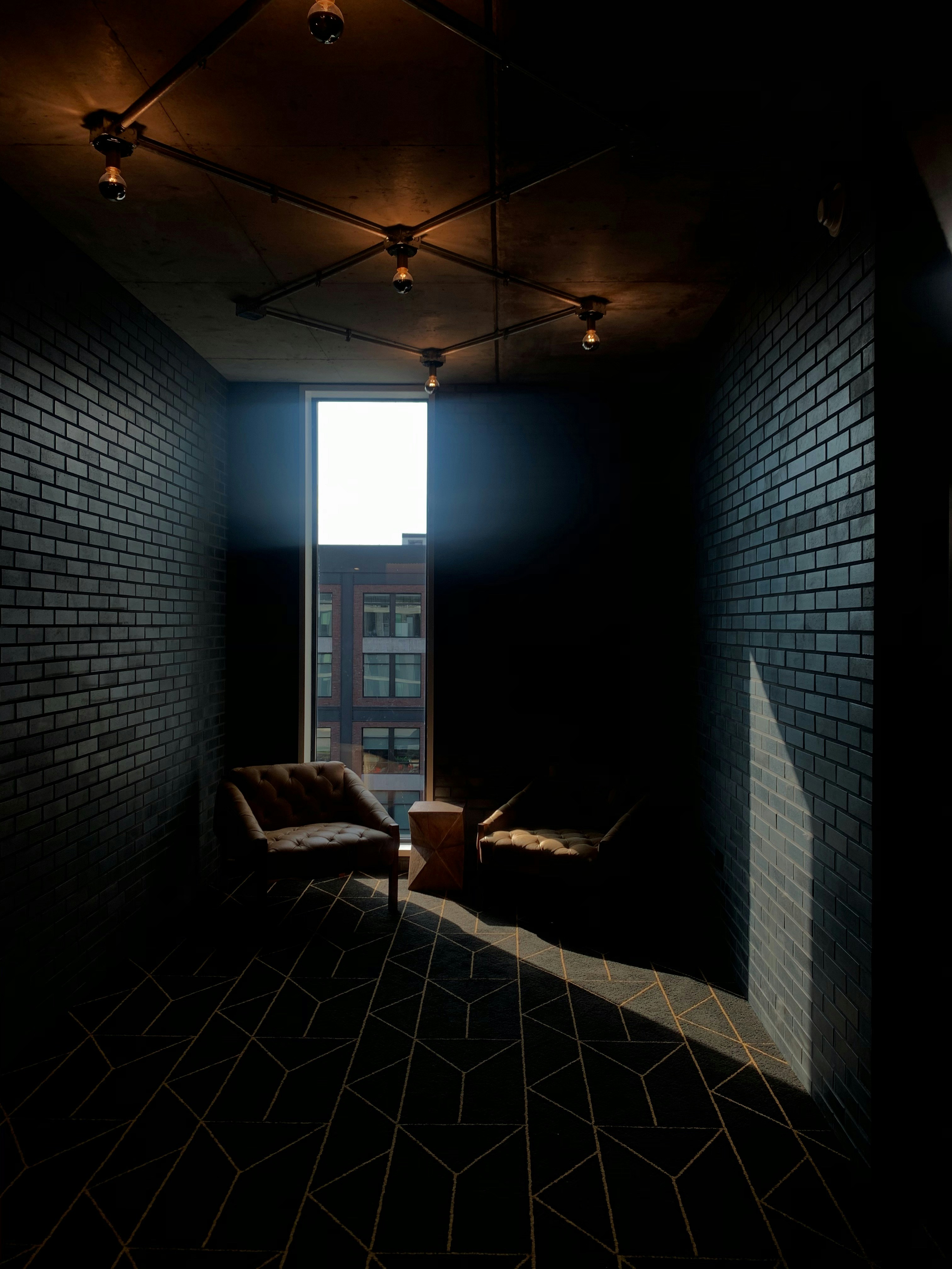 A cozy seating area illuminated by natural light filtering through a tall window, surrounded by dark brick walls and geometric flooring.