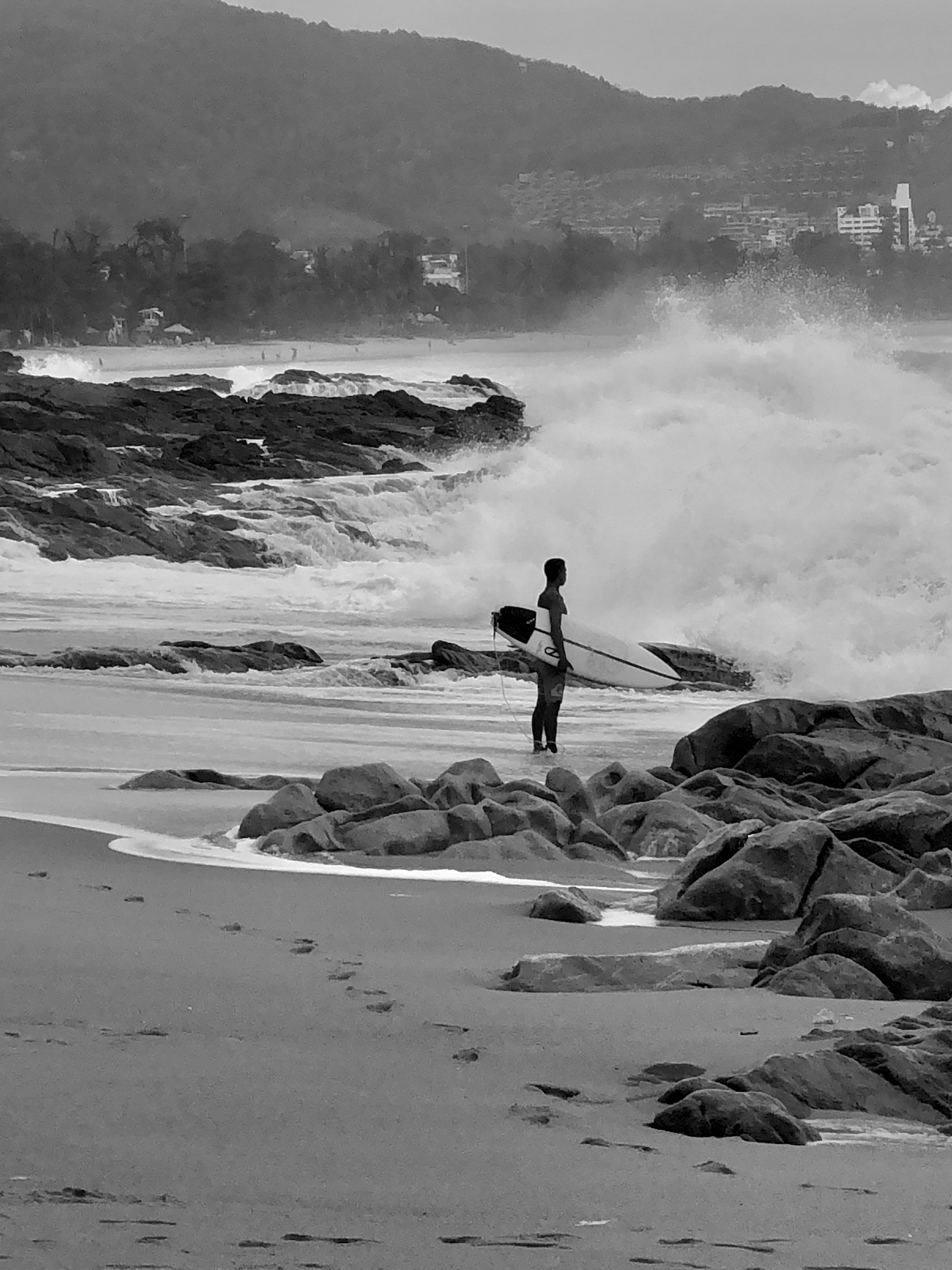 Surfer standing on rocky shoreline with crashing waves in the background on an overcast day.