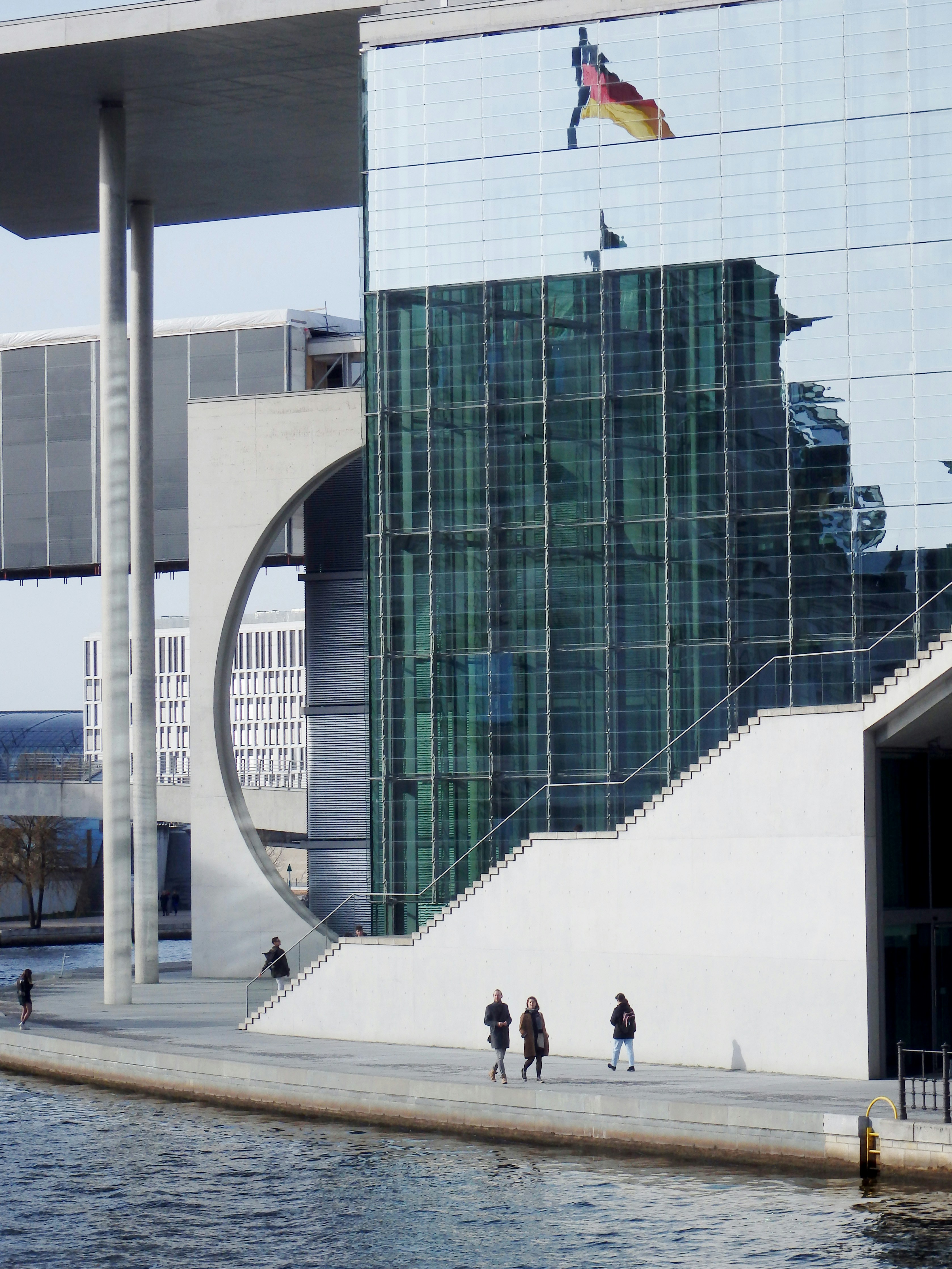 Modern glass building reflecting a German flag, with pedestrians walking along the waterfront promenade.