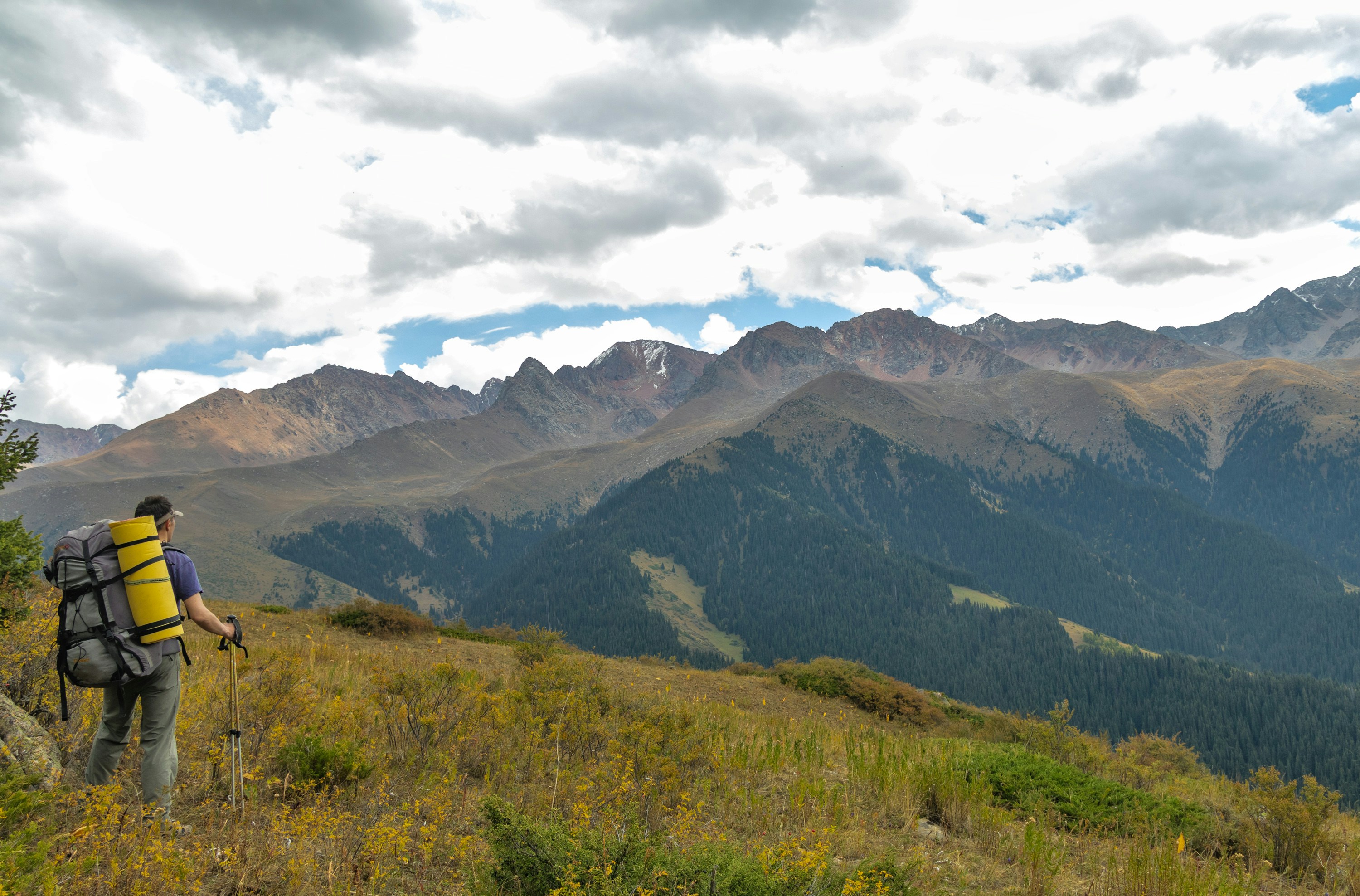 Hiker with a vibrant backpack overlooks a vast mountain range under a dramatic cloudy sky.