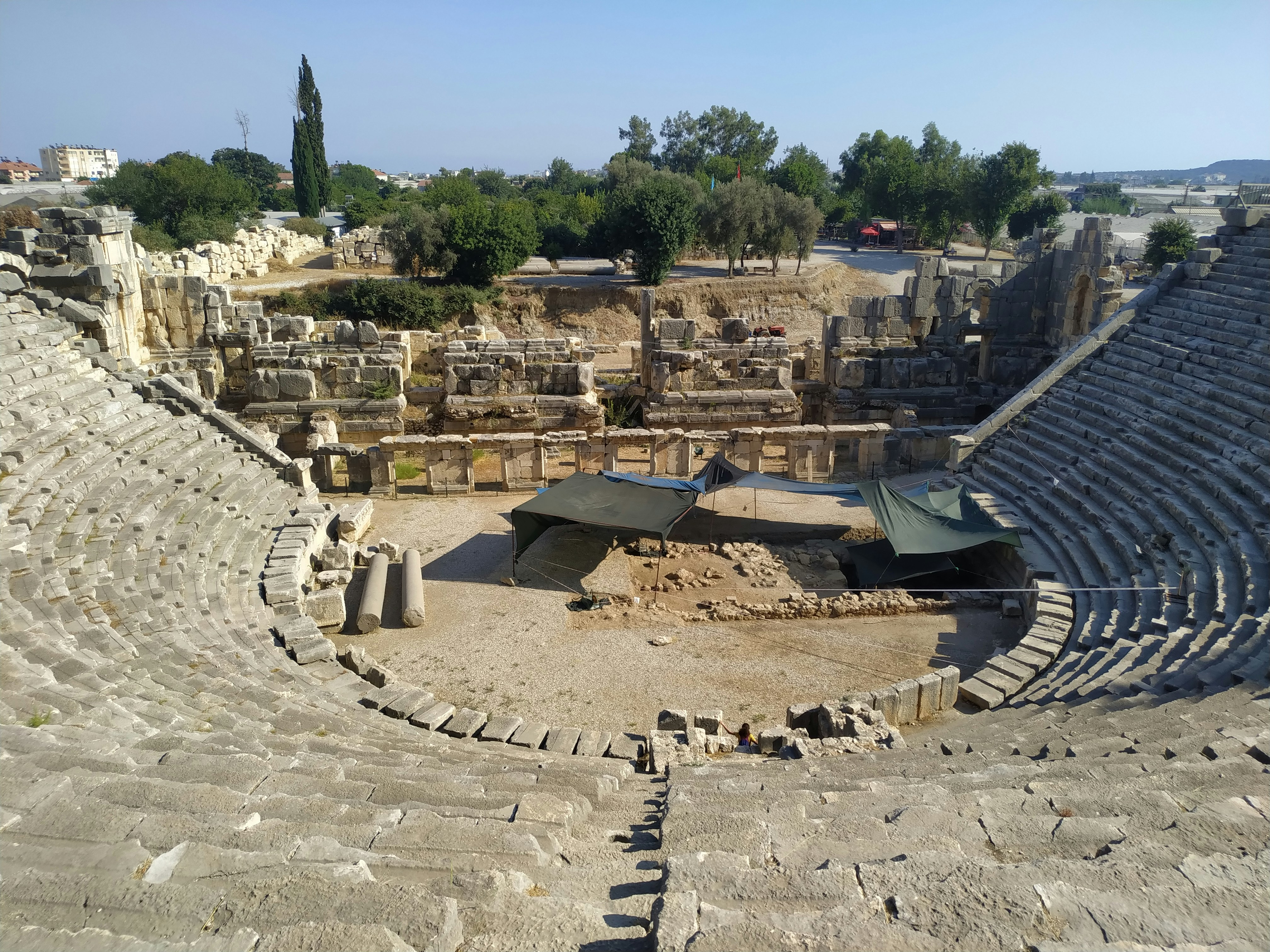 Ancient amphitheater showcasing stone seating and remnants of historical structures, with tents set up in the center. The scene captures the essence of bygone theatrical grandeur.